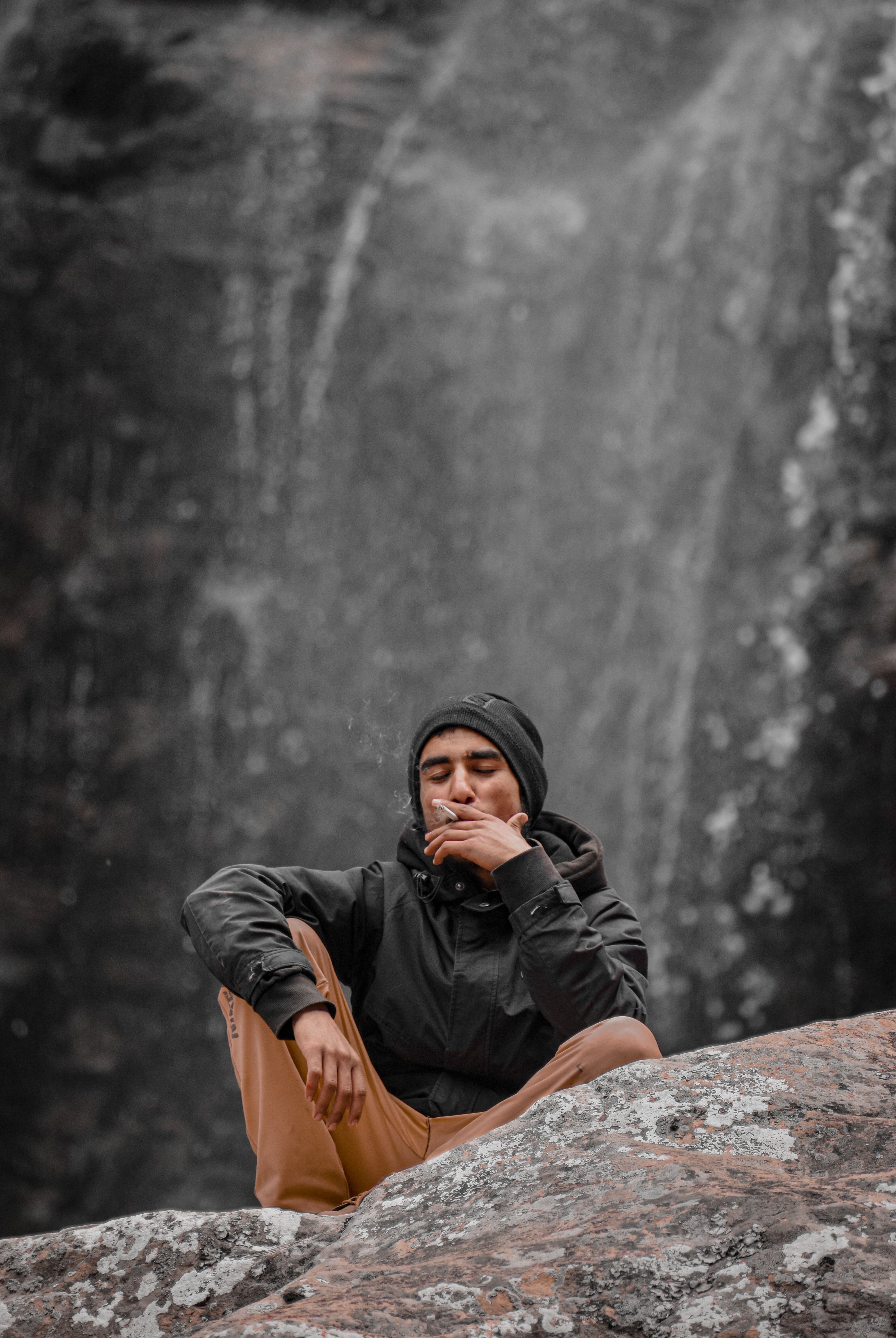 Man Crouching on a Stone with a Cigarette and Black Rock in Background ...