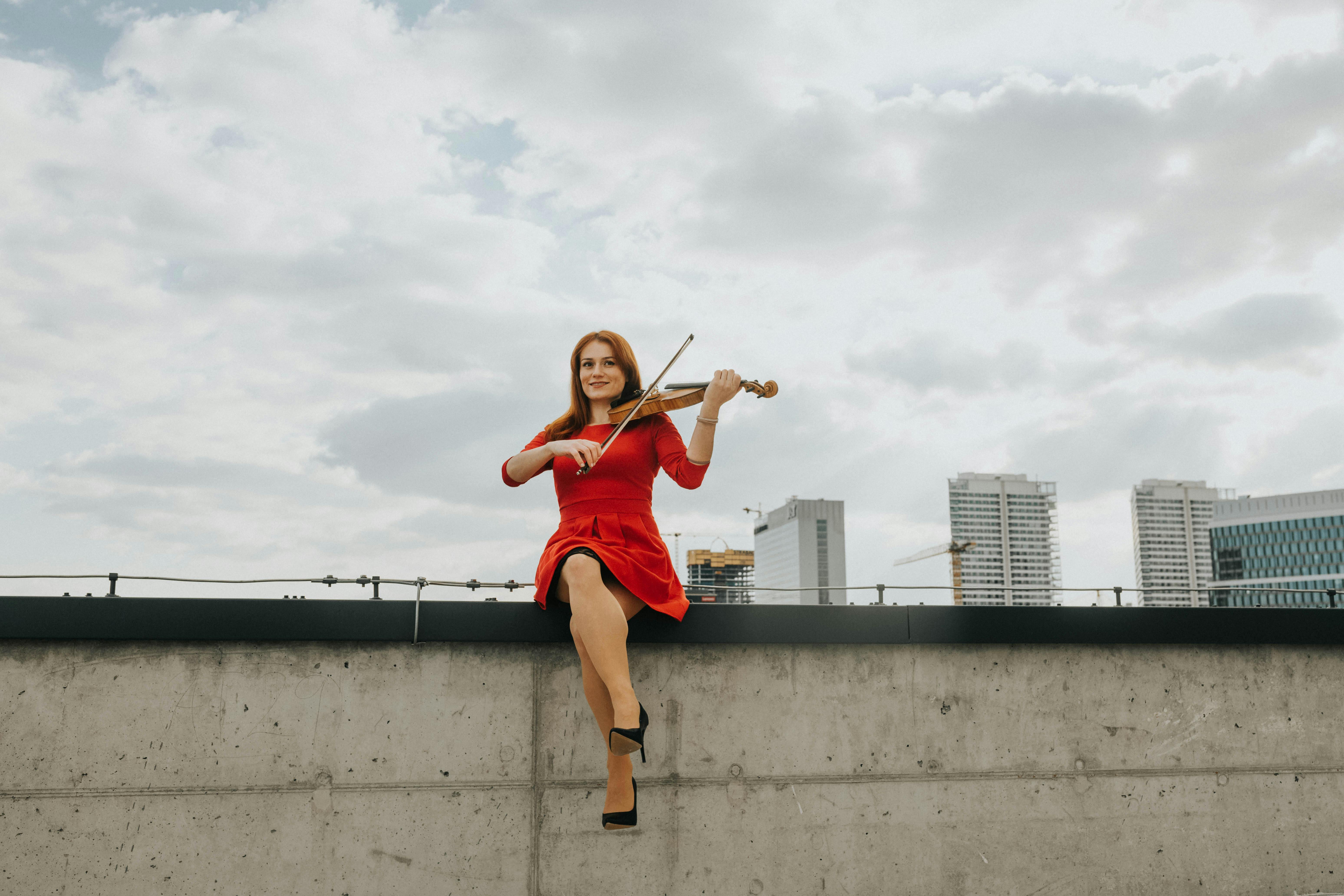 Woman in Red Dress Sitting on Concrete Fence While Playing Violin ...