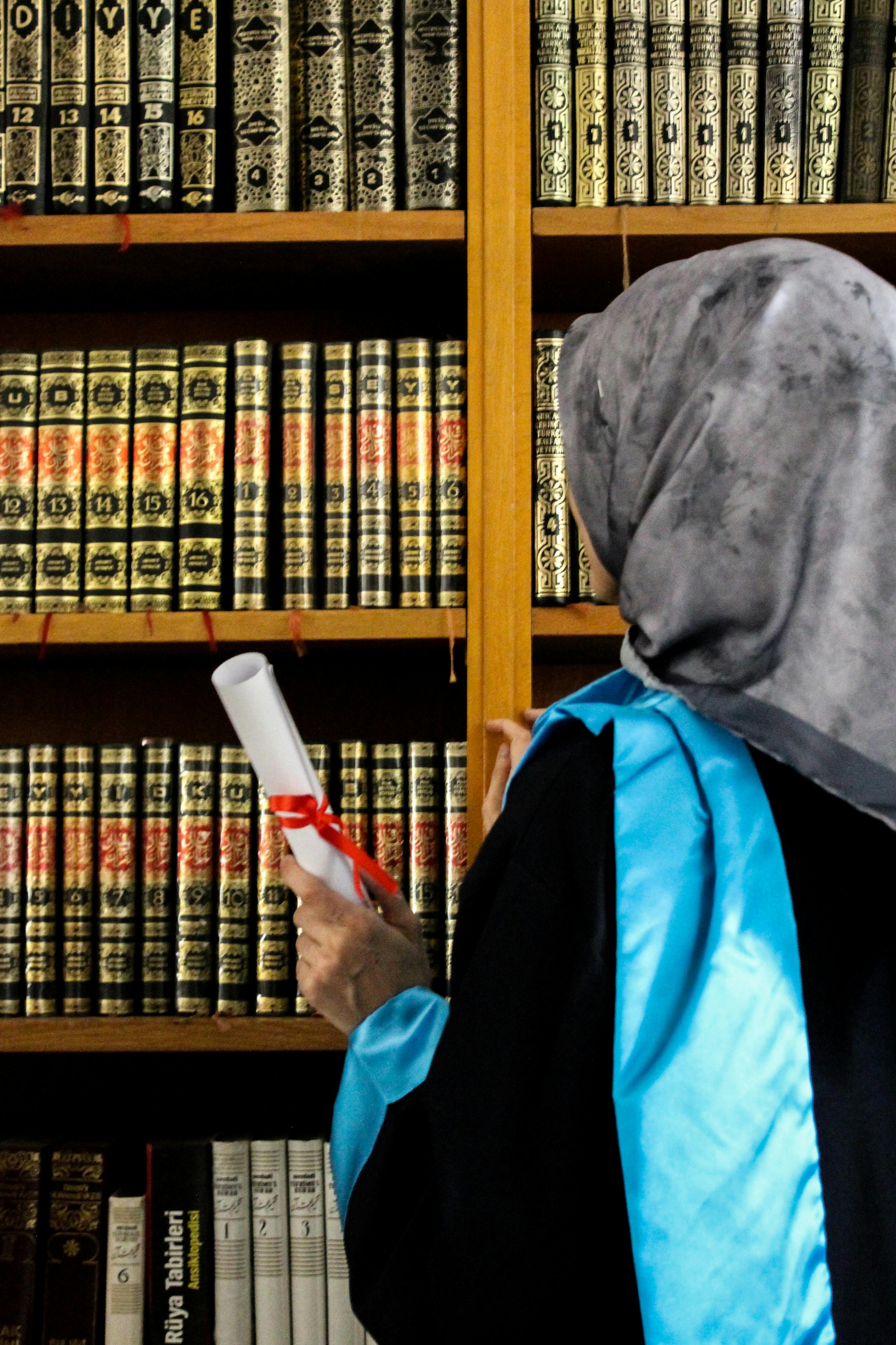 Hijabi Woman Holding Scroll Browsing Books in Library · Free Stock Photo