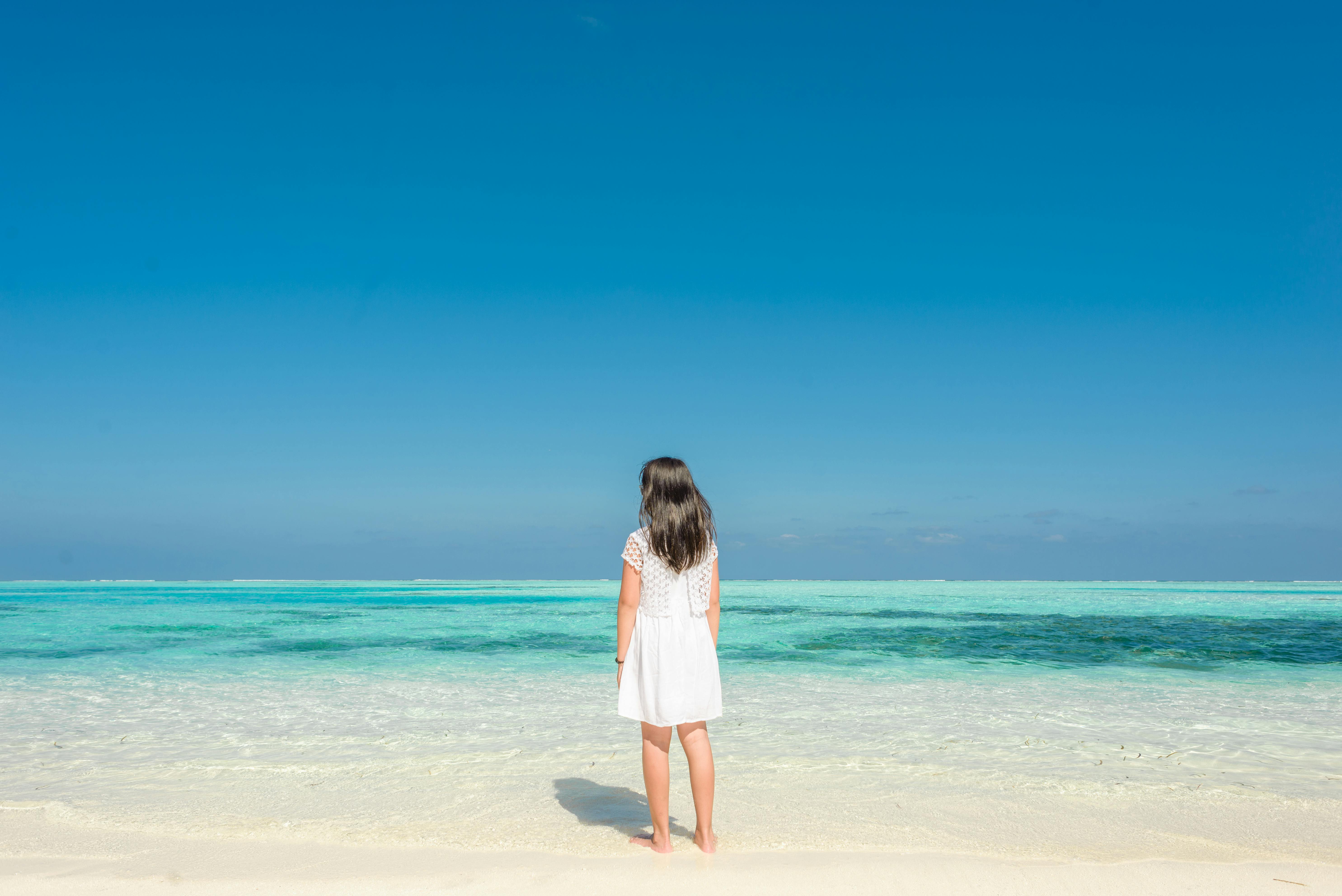 Back View of a Girl in white Dress Standing on the Beach under the Blue ...