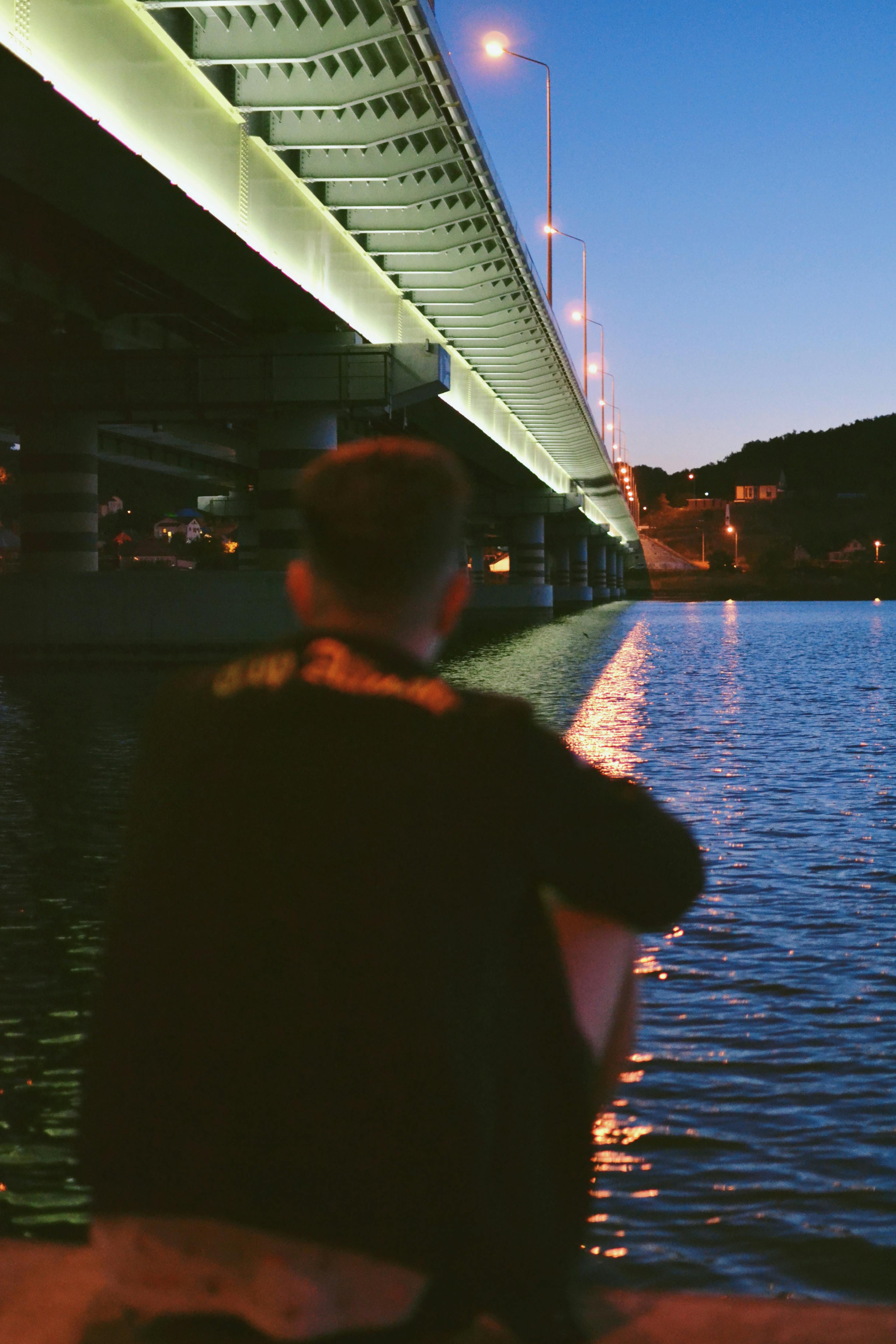 Man Sitting on River Shore under Bridge · Free Stock Photo