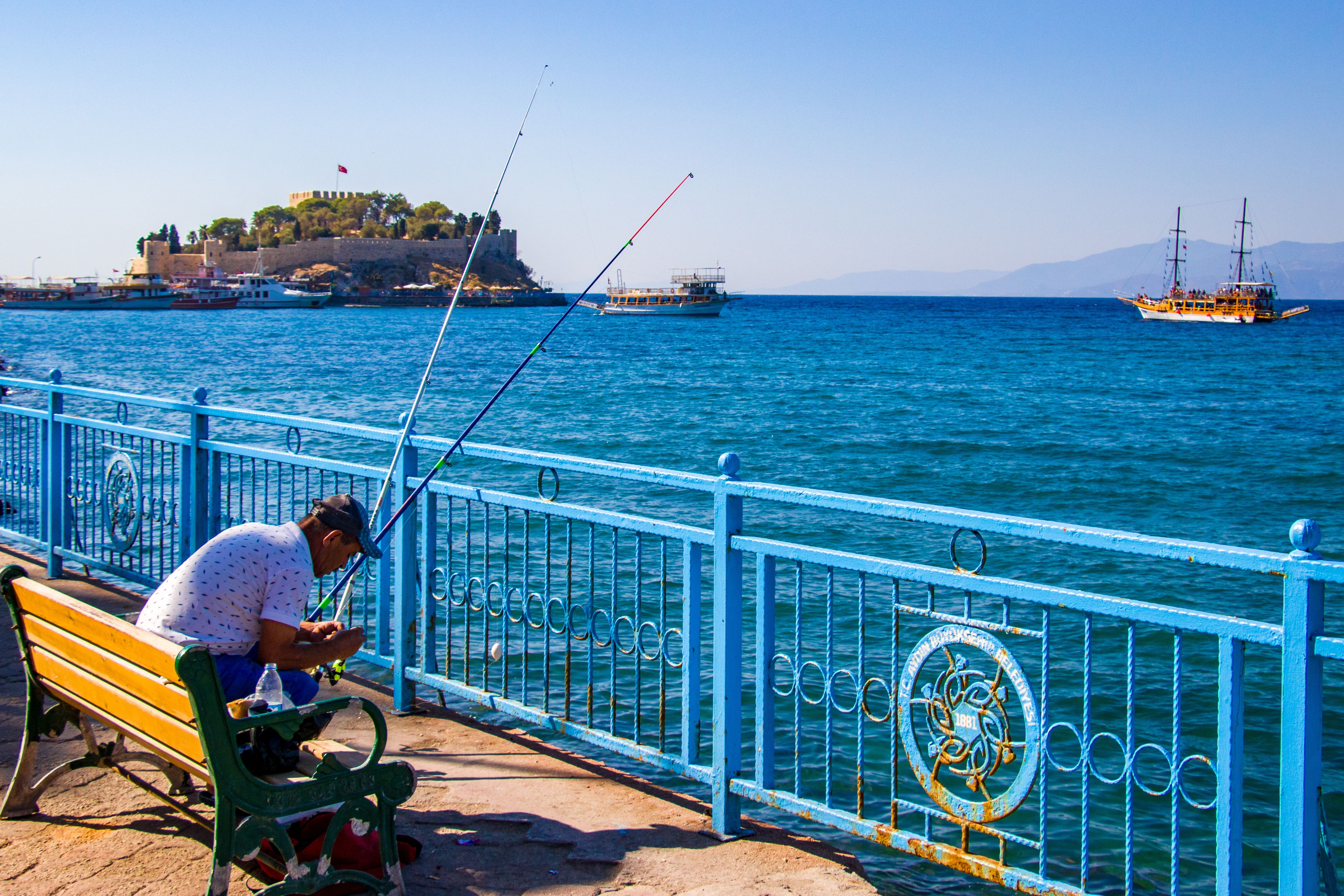 Man Sitting on a Bench Holding a Fishing Rod Near a Body of Water ...