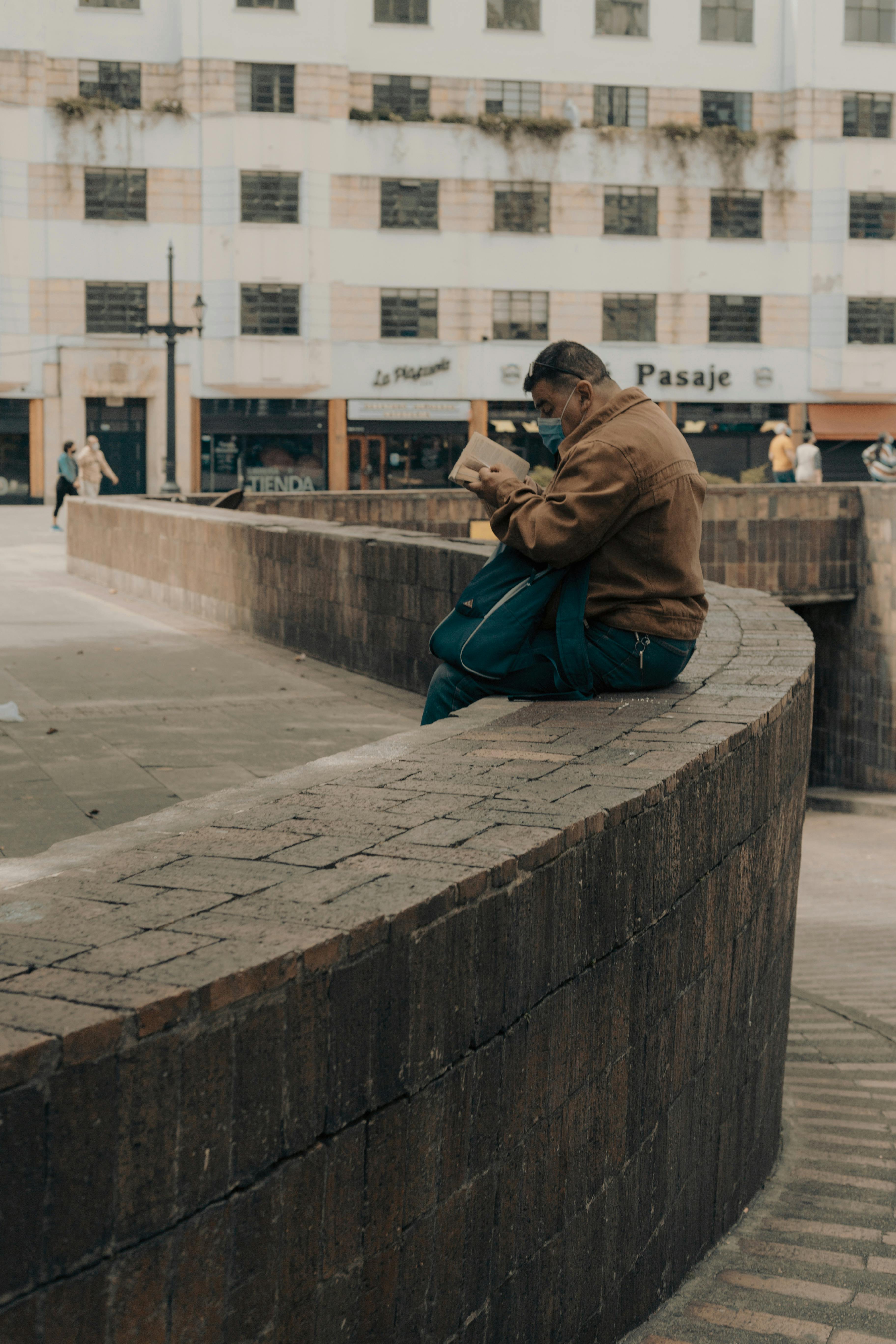 Man Sitting on a Brown Wall and Reading a Book in a Town Square · Free ...