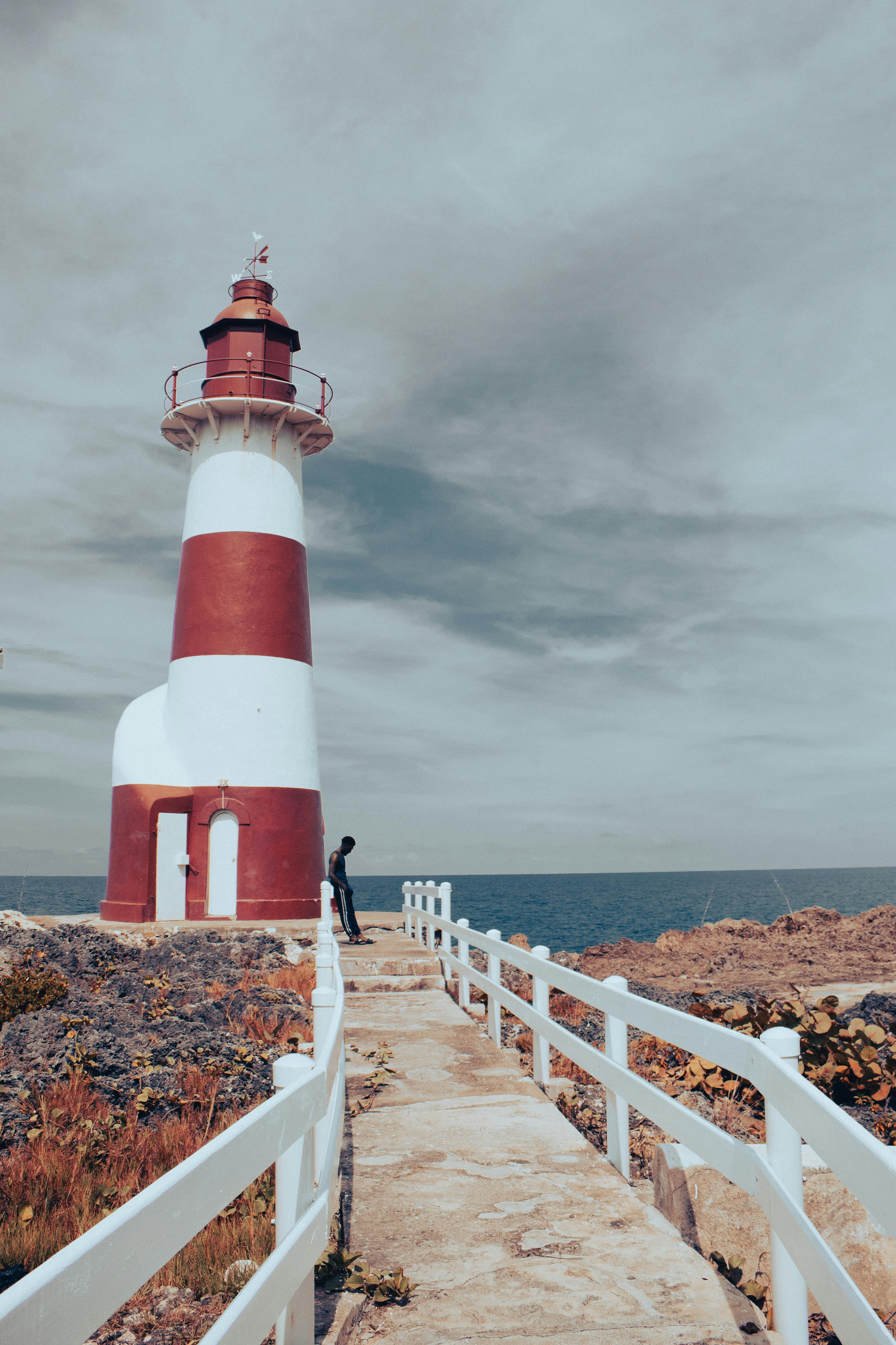 A Person Leaning on the Wooden Railing Near White and Red Lighthouse ...