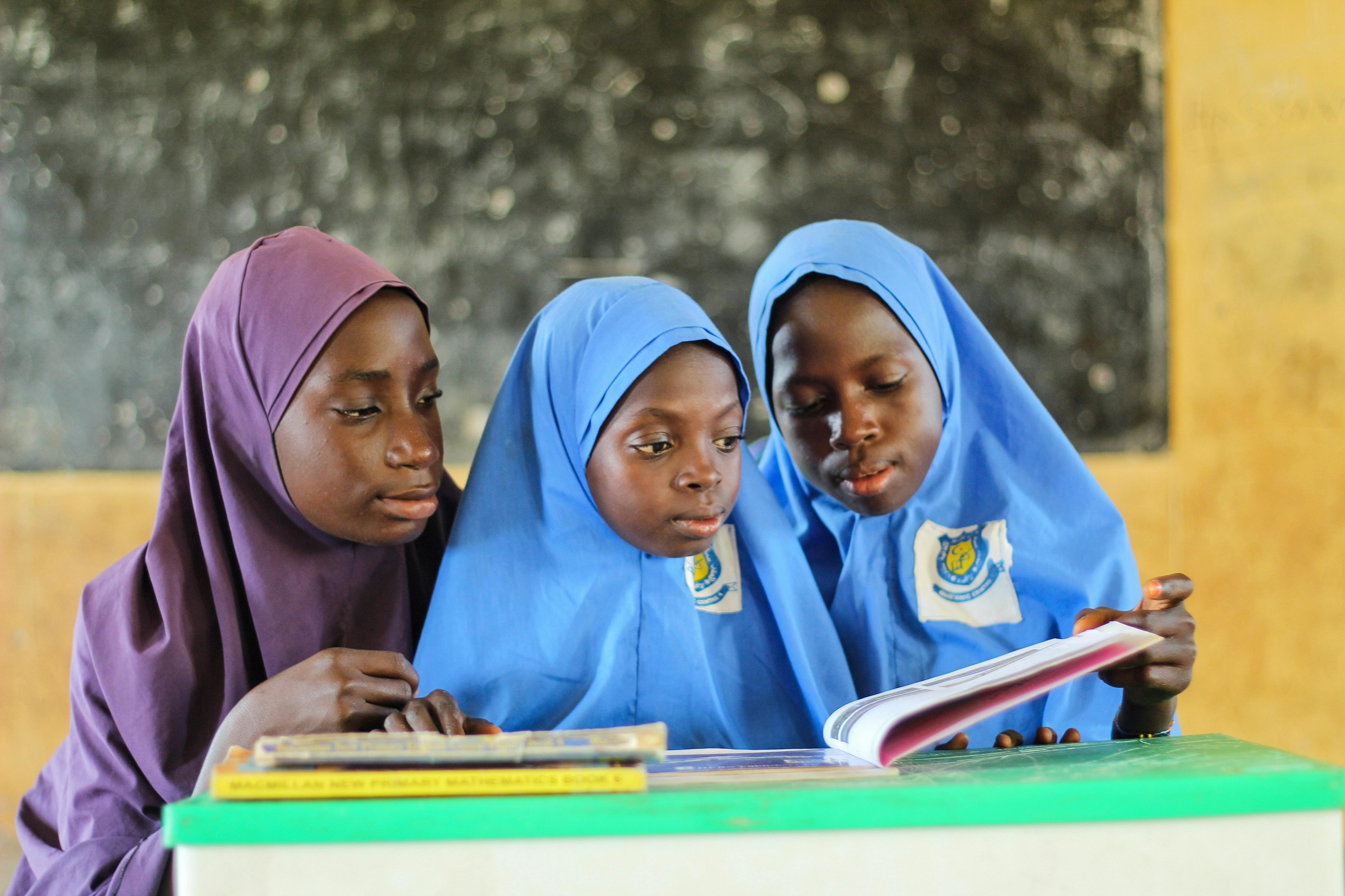 A Pair of Girls Wearing Blue Hijabs Reading a Book Beside a Young Woman ...