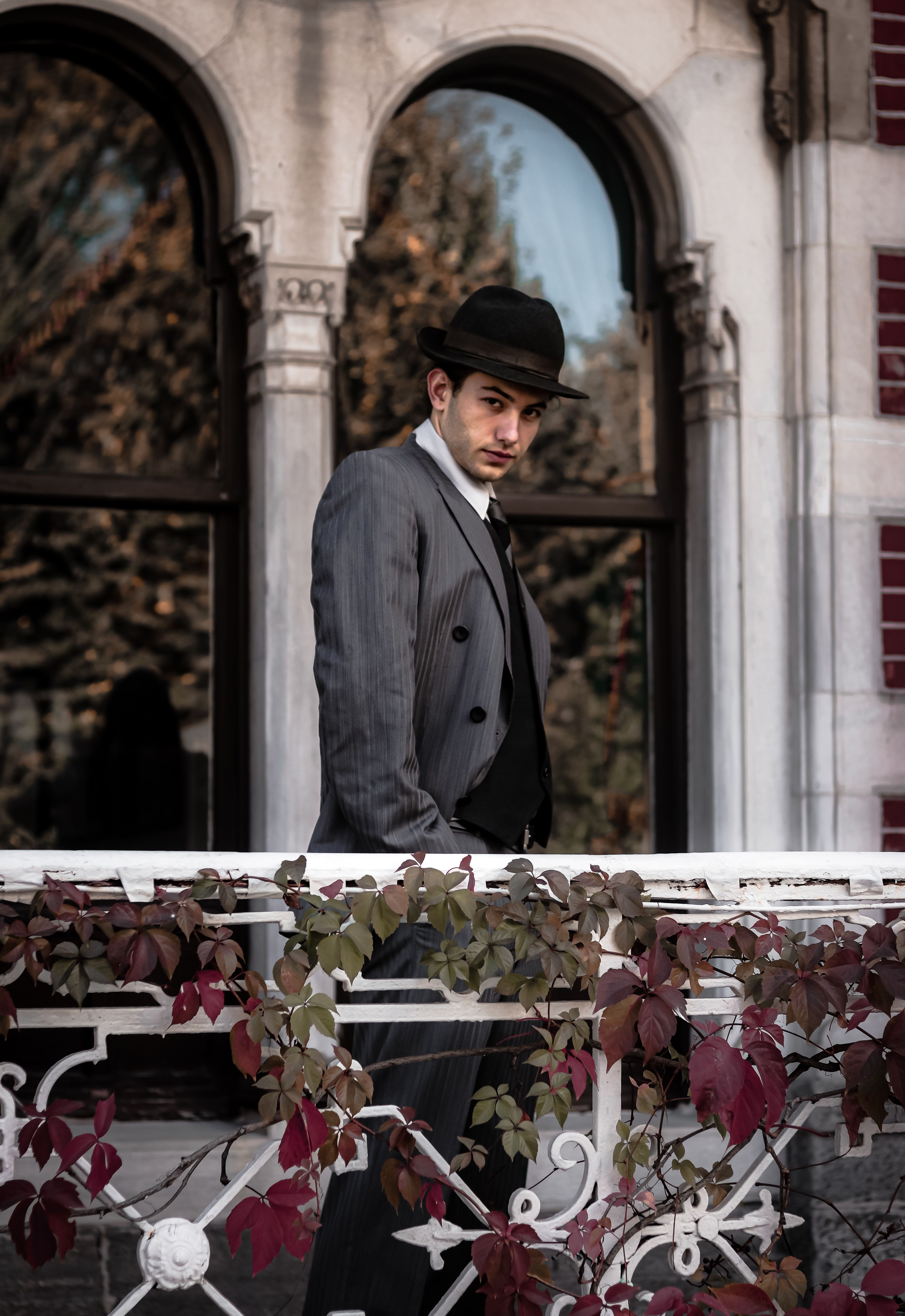 Elegant Man in Hat Standing on Balcony Covered in Vines · Free Stock Photo