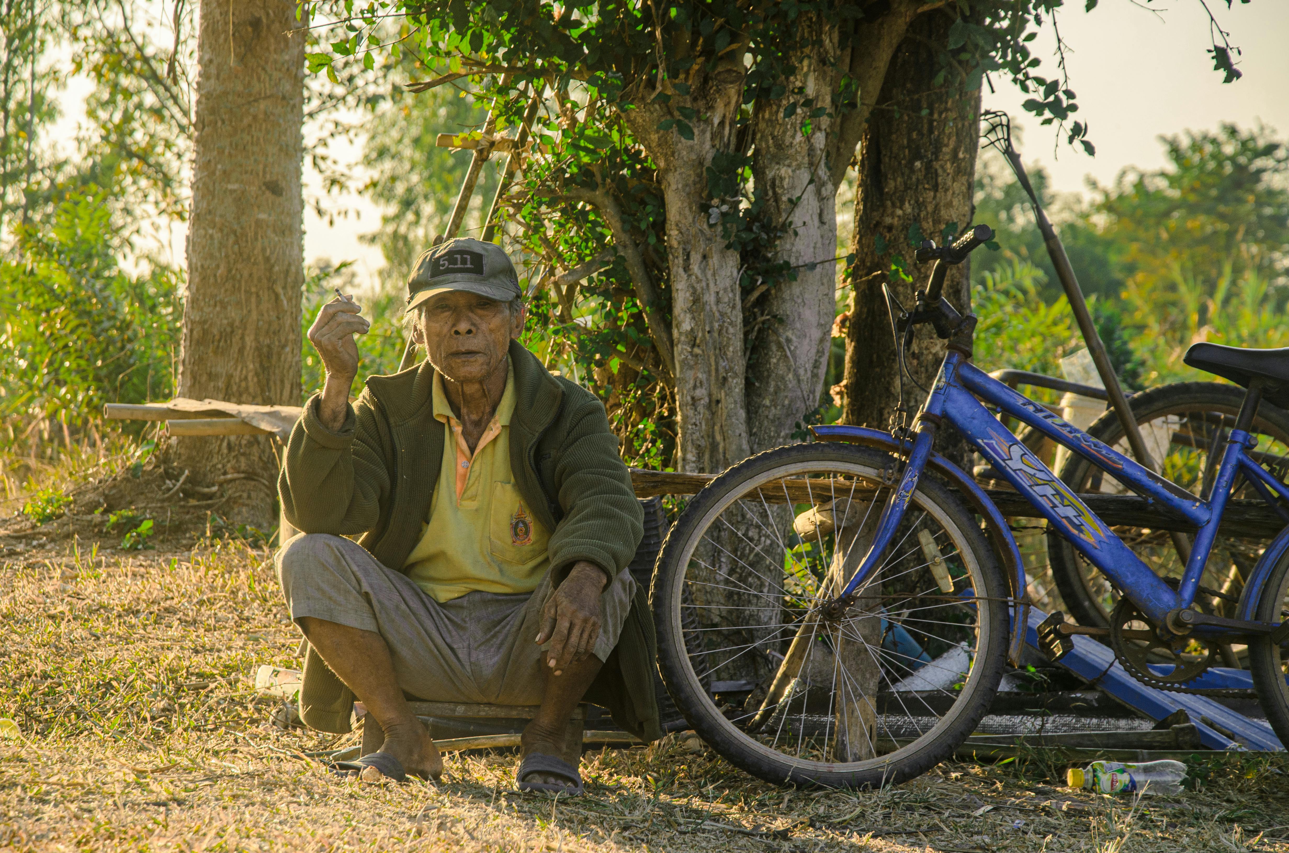 Senior Man Crouching by a Blue Bicycle under a Tree, and Smoking a ...