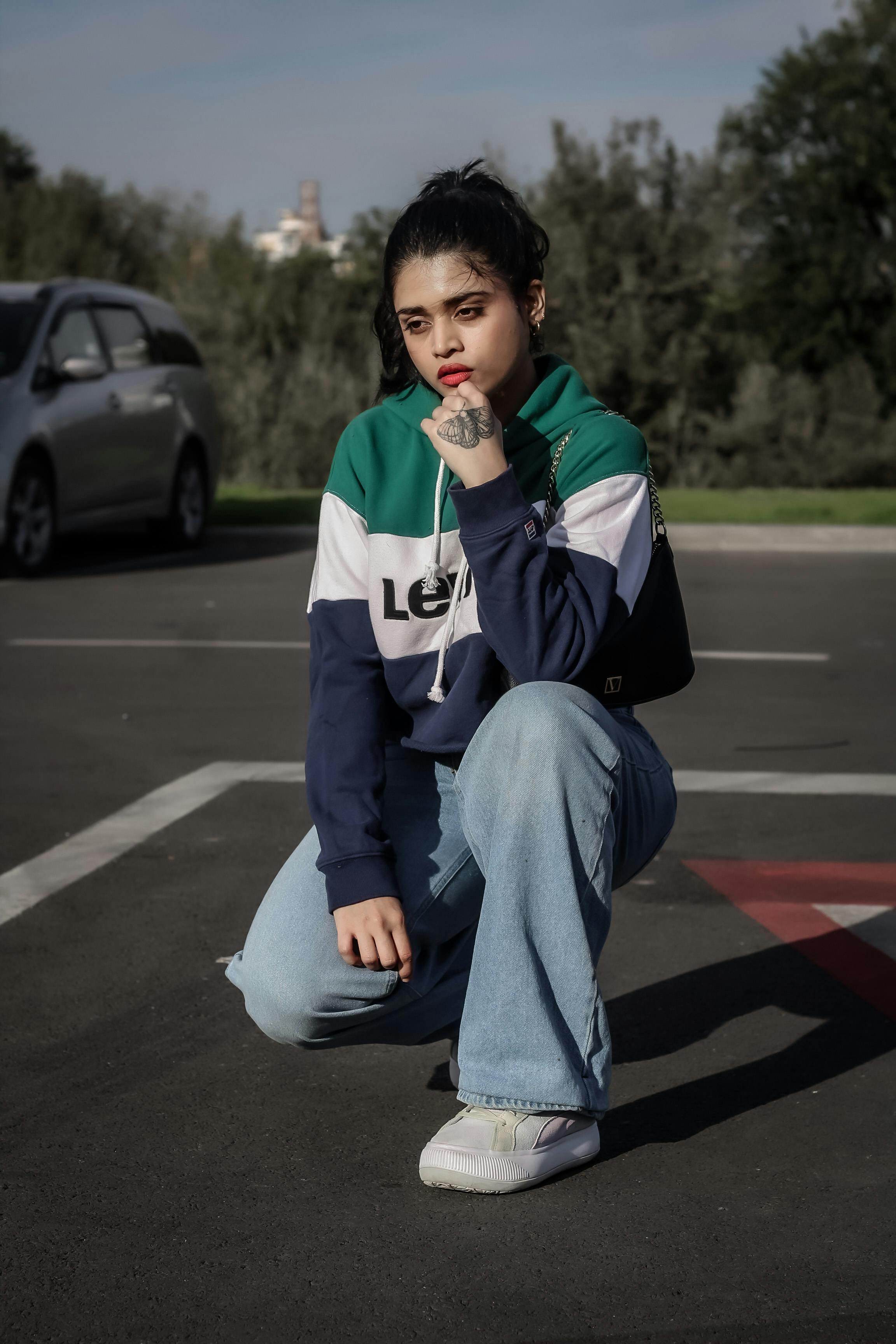 Young Woman in Hoodie and Jeans Crouching on Parking Lot · Free Stock Photo
