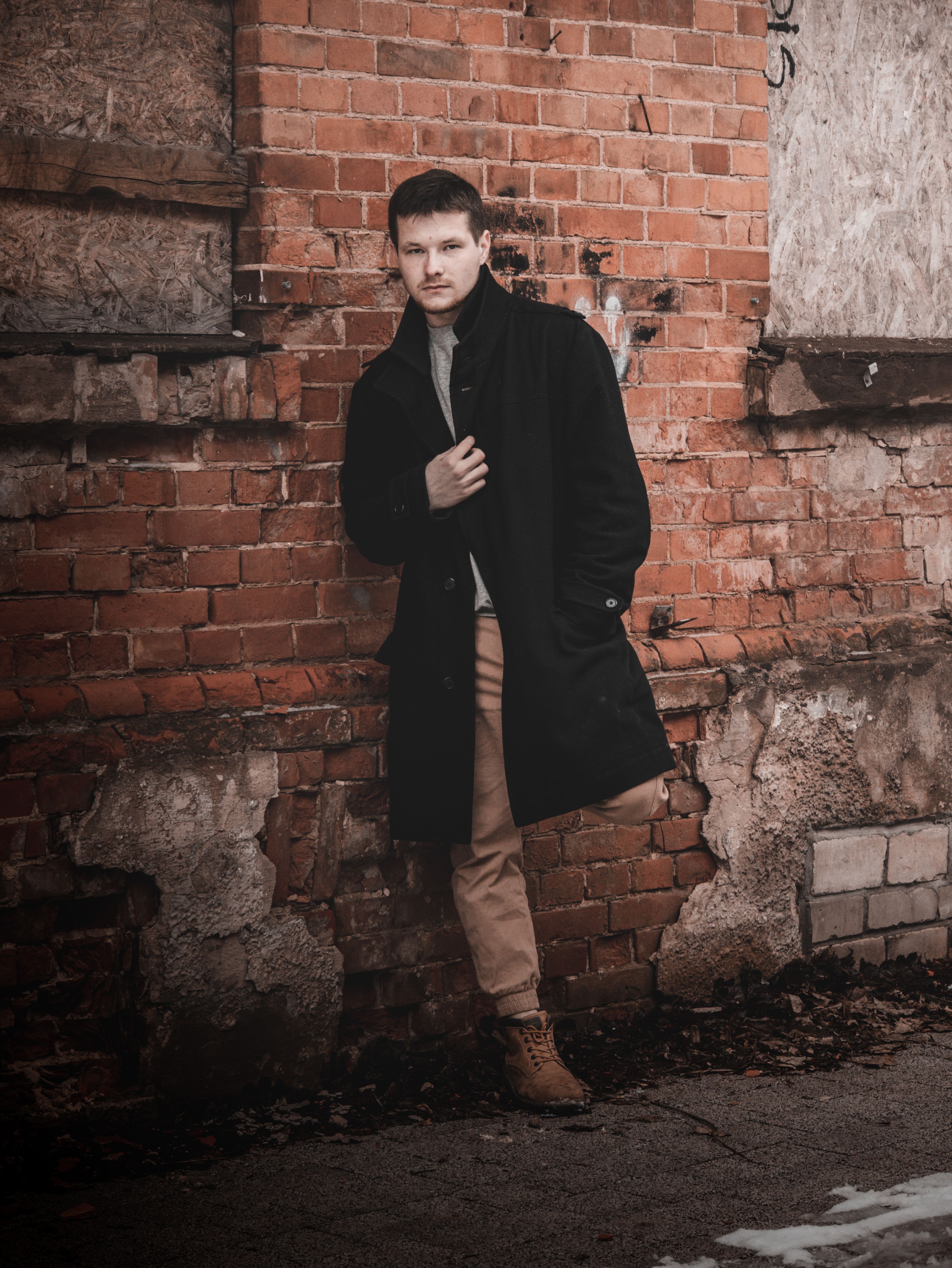 Male Model Wearing a Black Coat Standing on One Leg in Front of a Brick ...