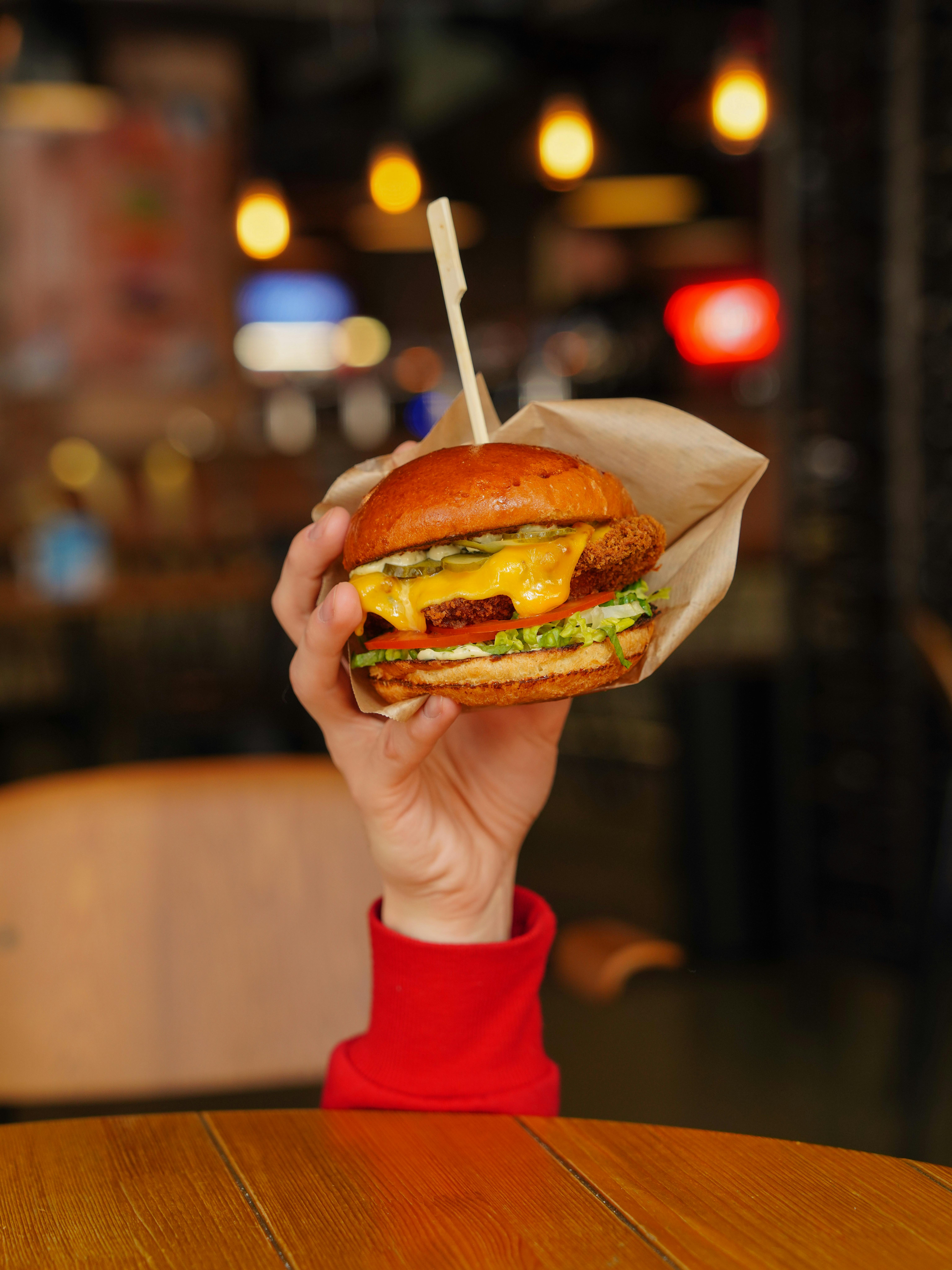 Hand Holding Cheeseburger over Table · Free Stock Photo