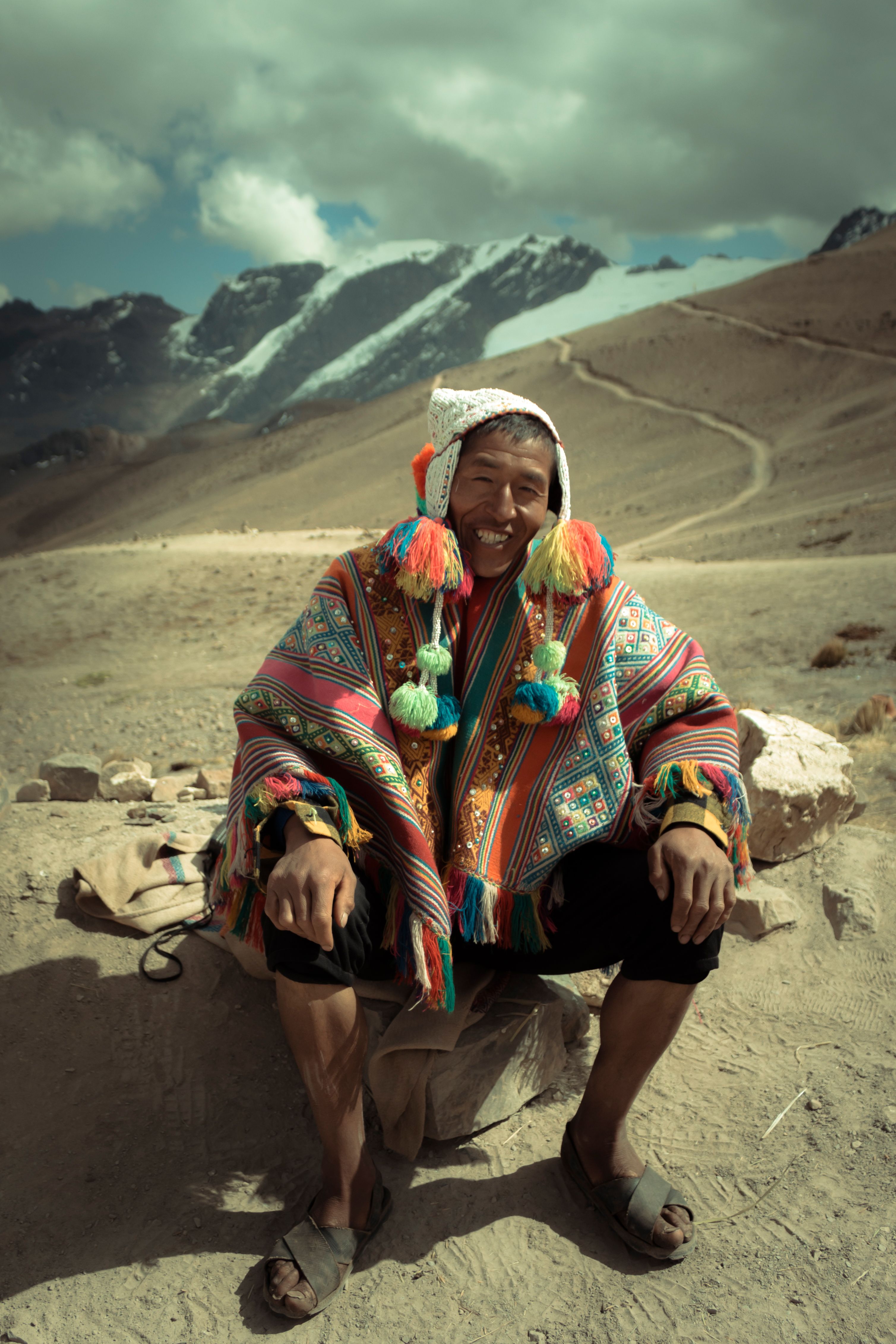 Smiling Man in Poncho Sitting and Posing in Mountains · Free Stock Photo