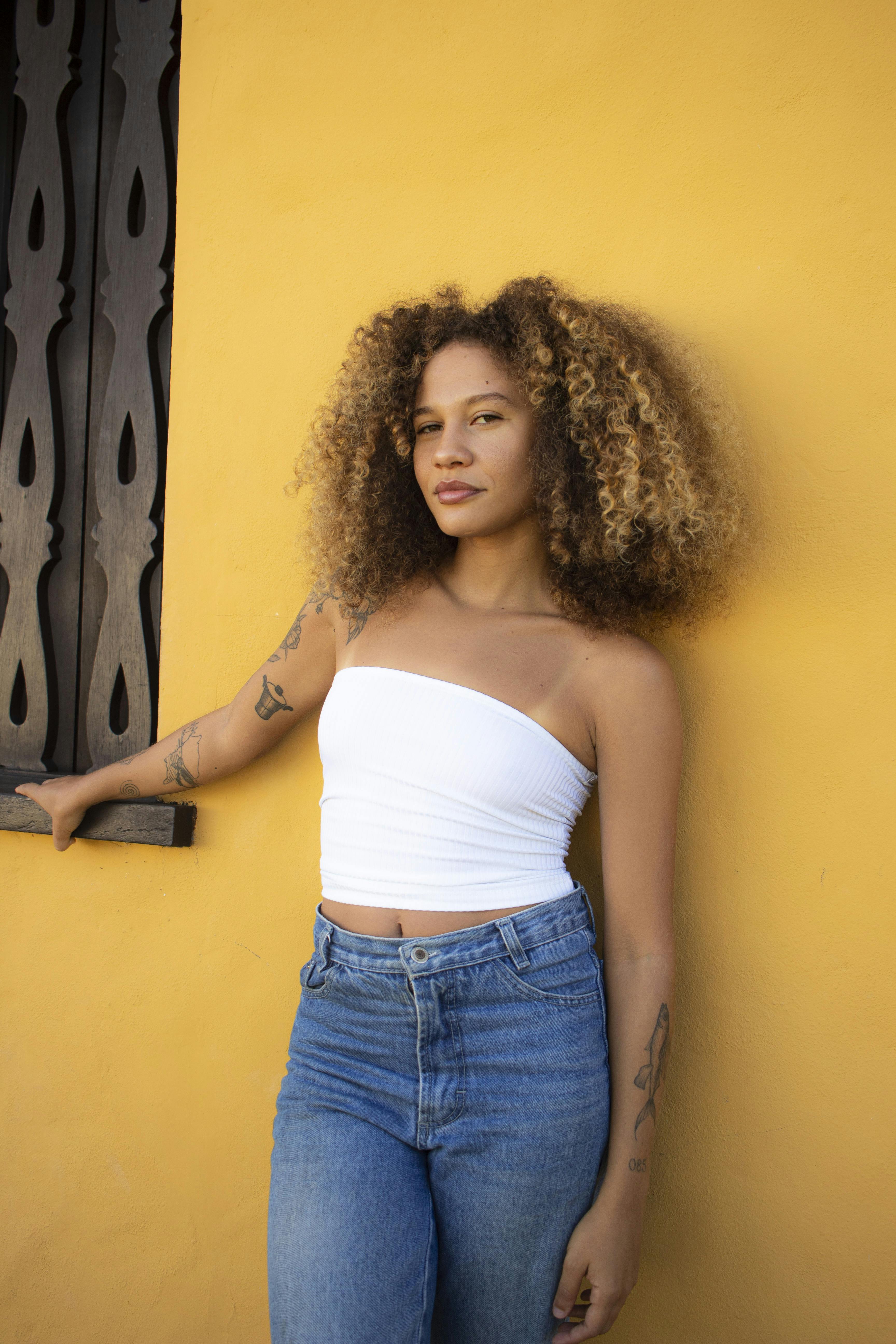 Young Woman in a White Top and Jeans Standing in front of a Yellow Wall · Free Stock Photo