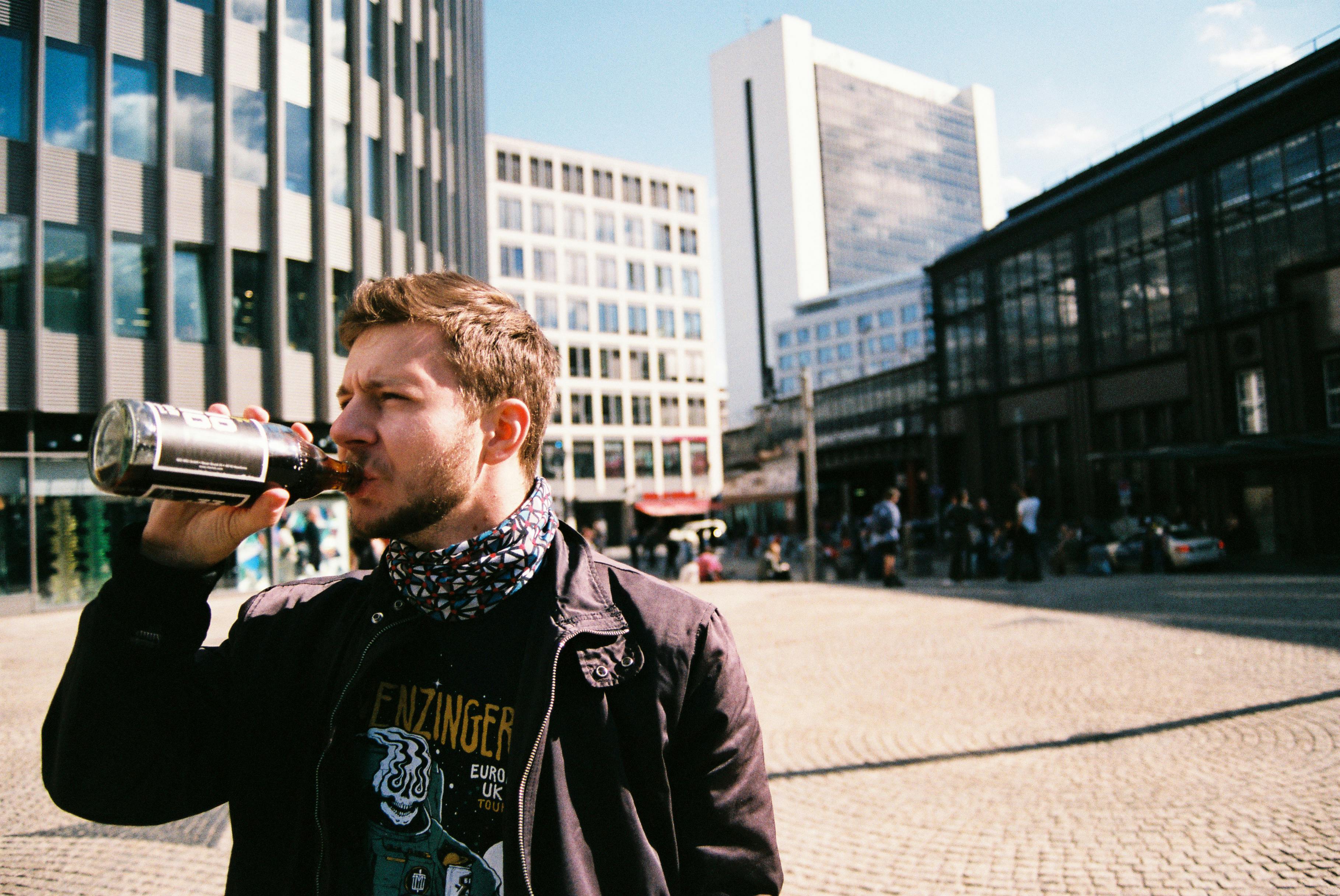 Man Drinking from Glass Bottle Standing on Plaza between Buildings ...