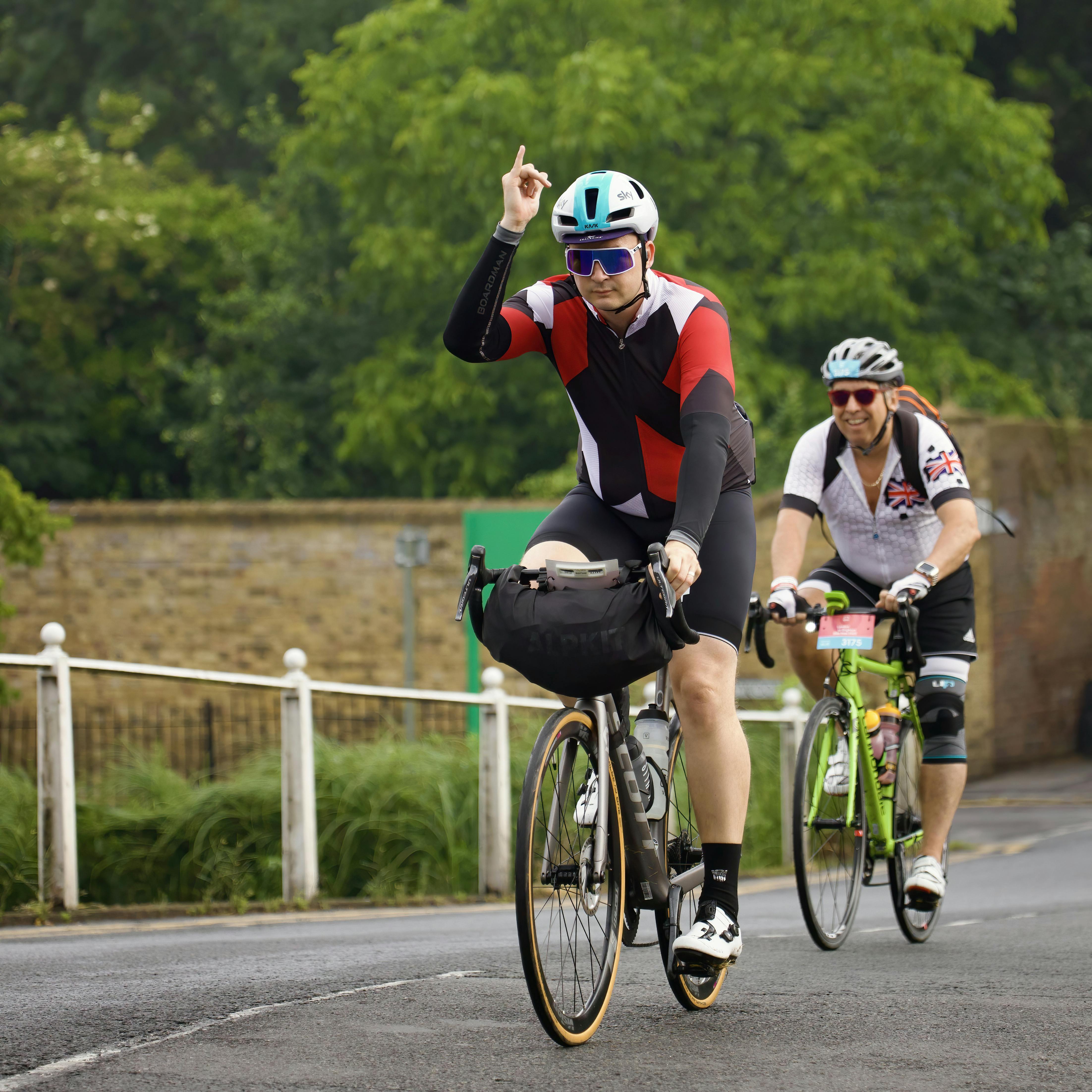 Two cyclists are riding down a road with one waving · Free Stock Photo