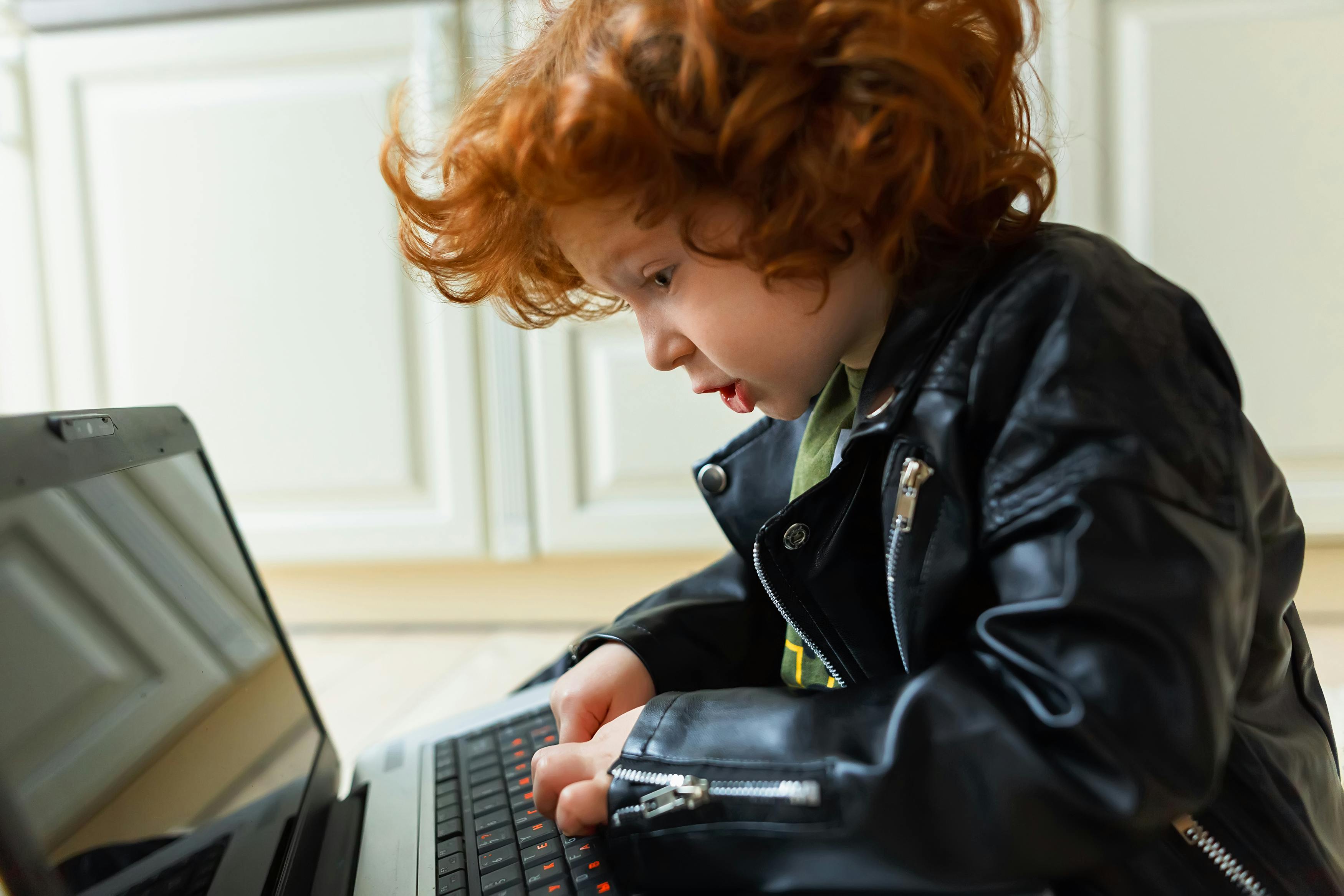 Little redhead boy uses a laptop while sitting on the floor at home ...