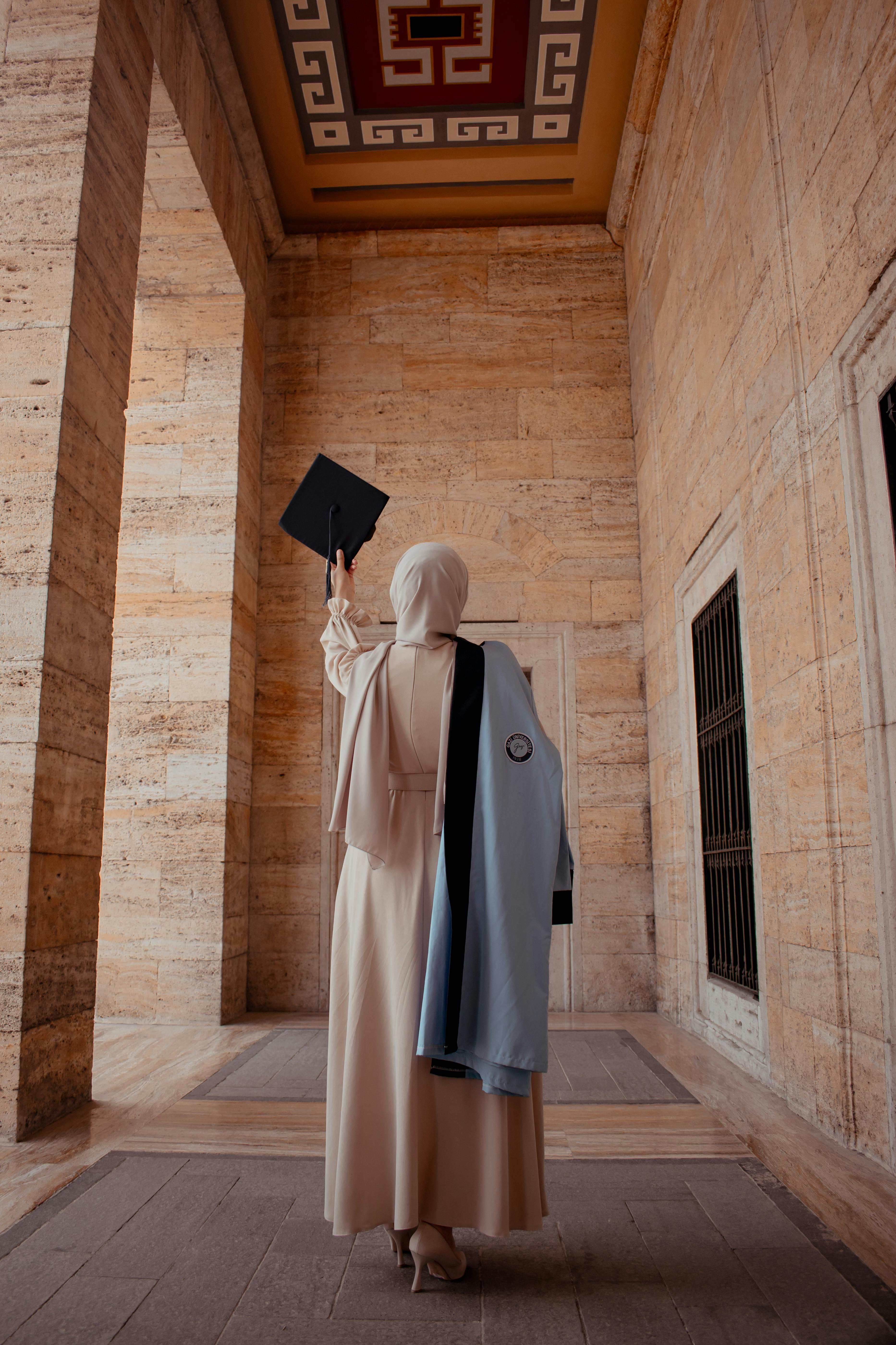 Graduate in Hijab and Gown Holding Academic Hat near Walls of Anitkabir ...