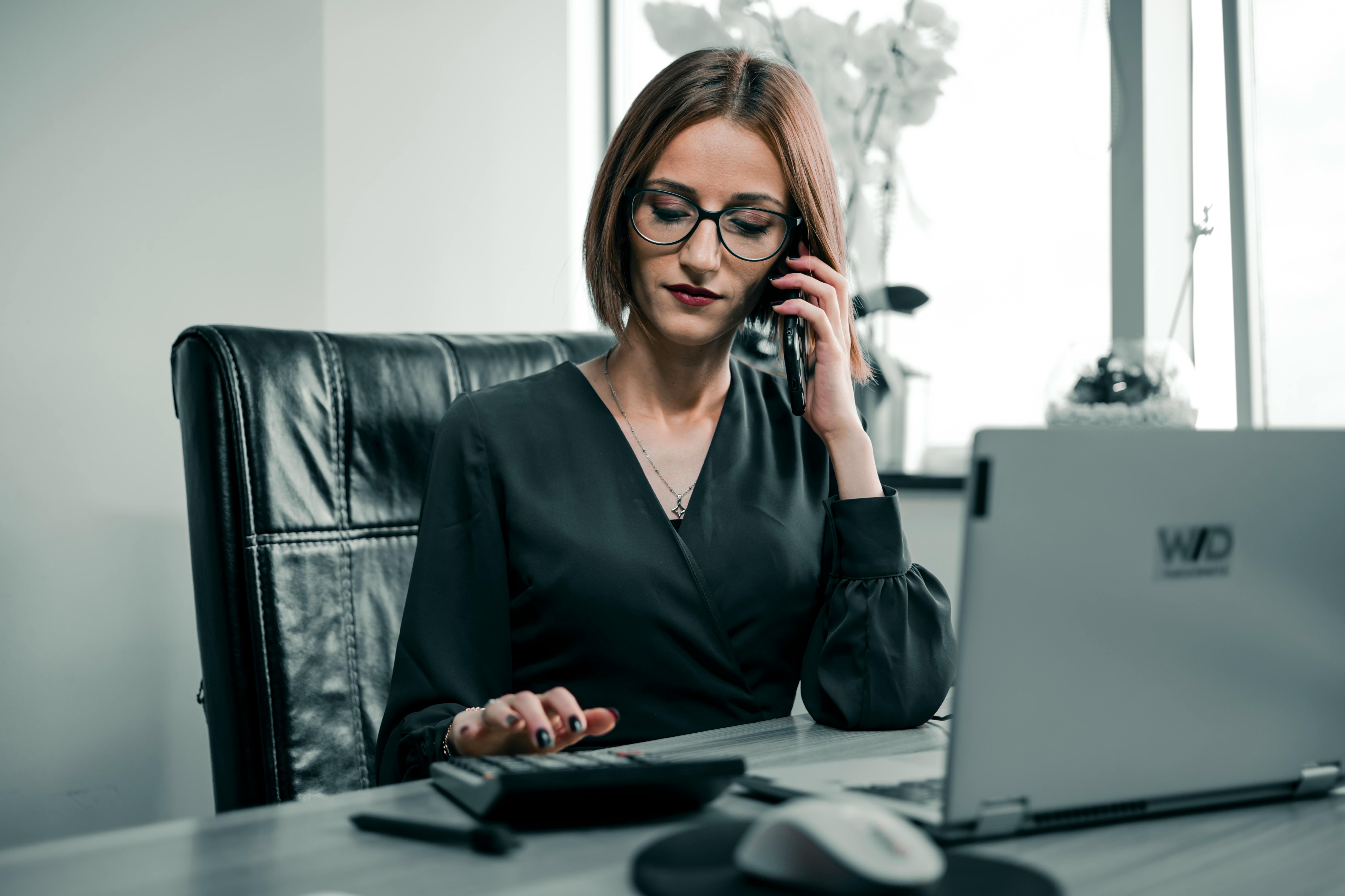 Woman Sitting by Office Desk Taking Call while Using Calcul · Free ...