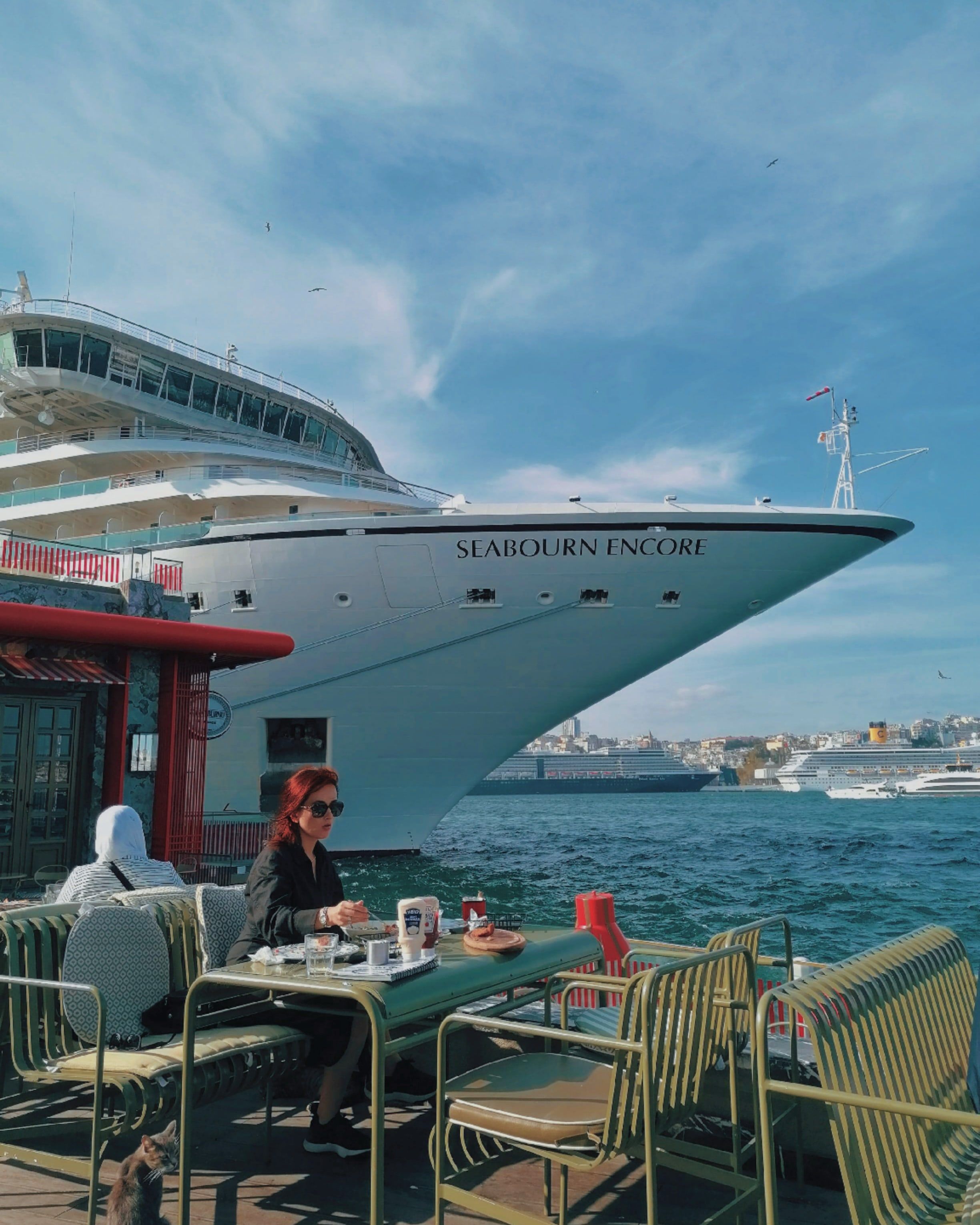 Woman Sitting by Table on Shore with Cruise Ship behind · Free Stock Photo