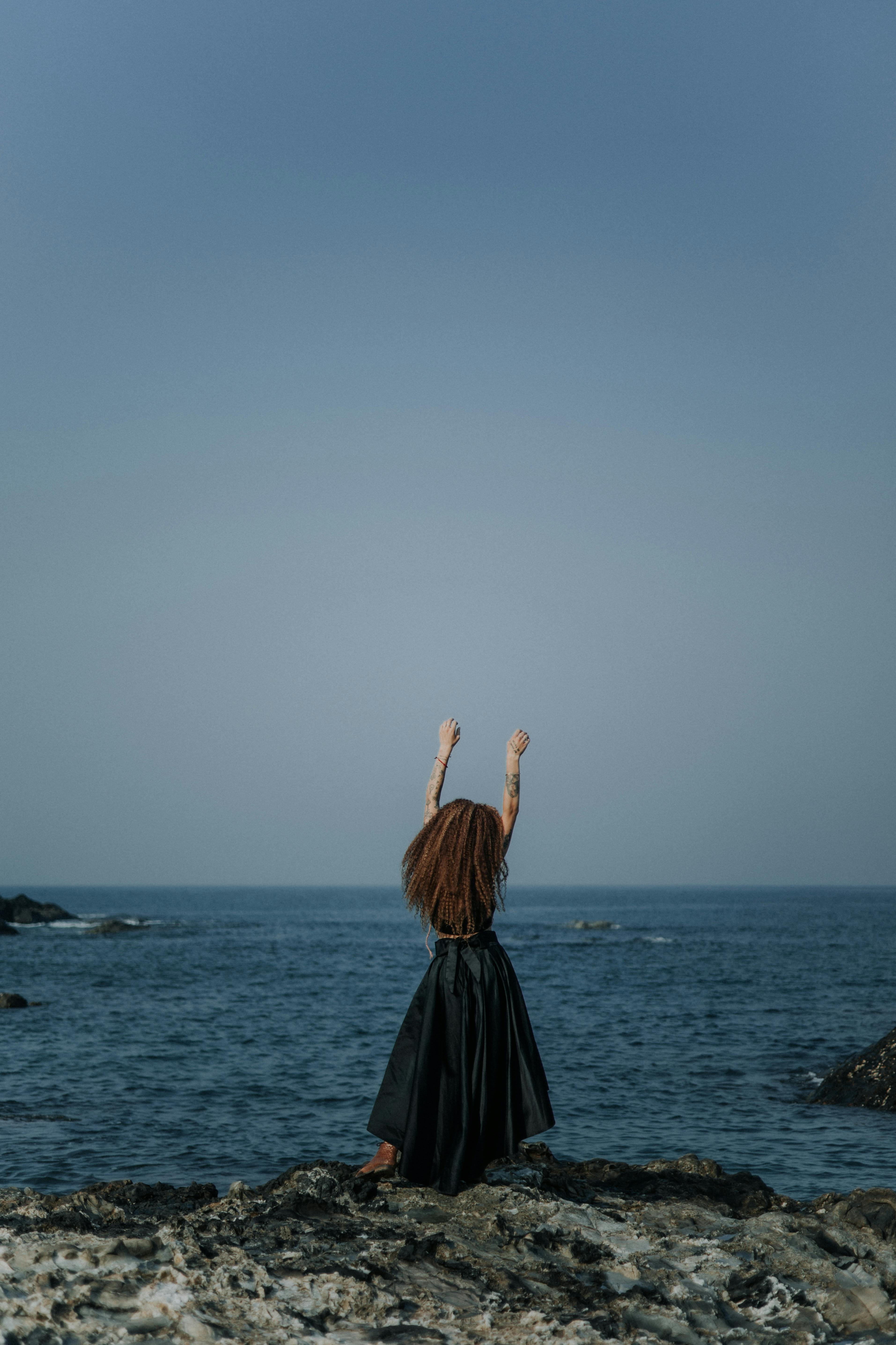 Back View of a Woman in a Black Dress Standing on a Shore with Arms ...
