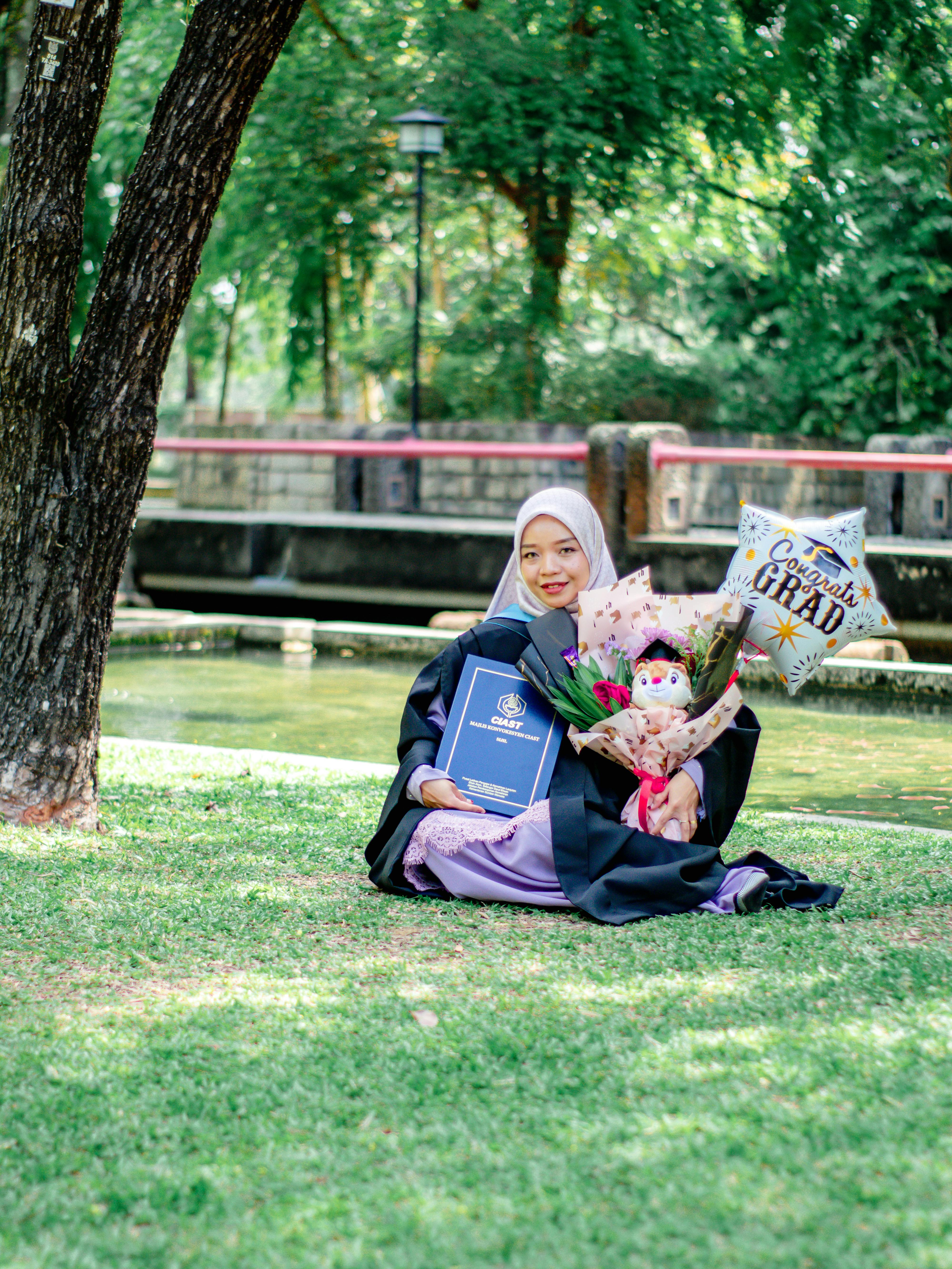A Girl in a Graduation Gown Sitting on the Grass in a Park with Flowers ...