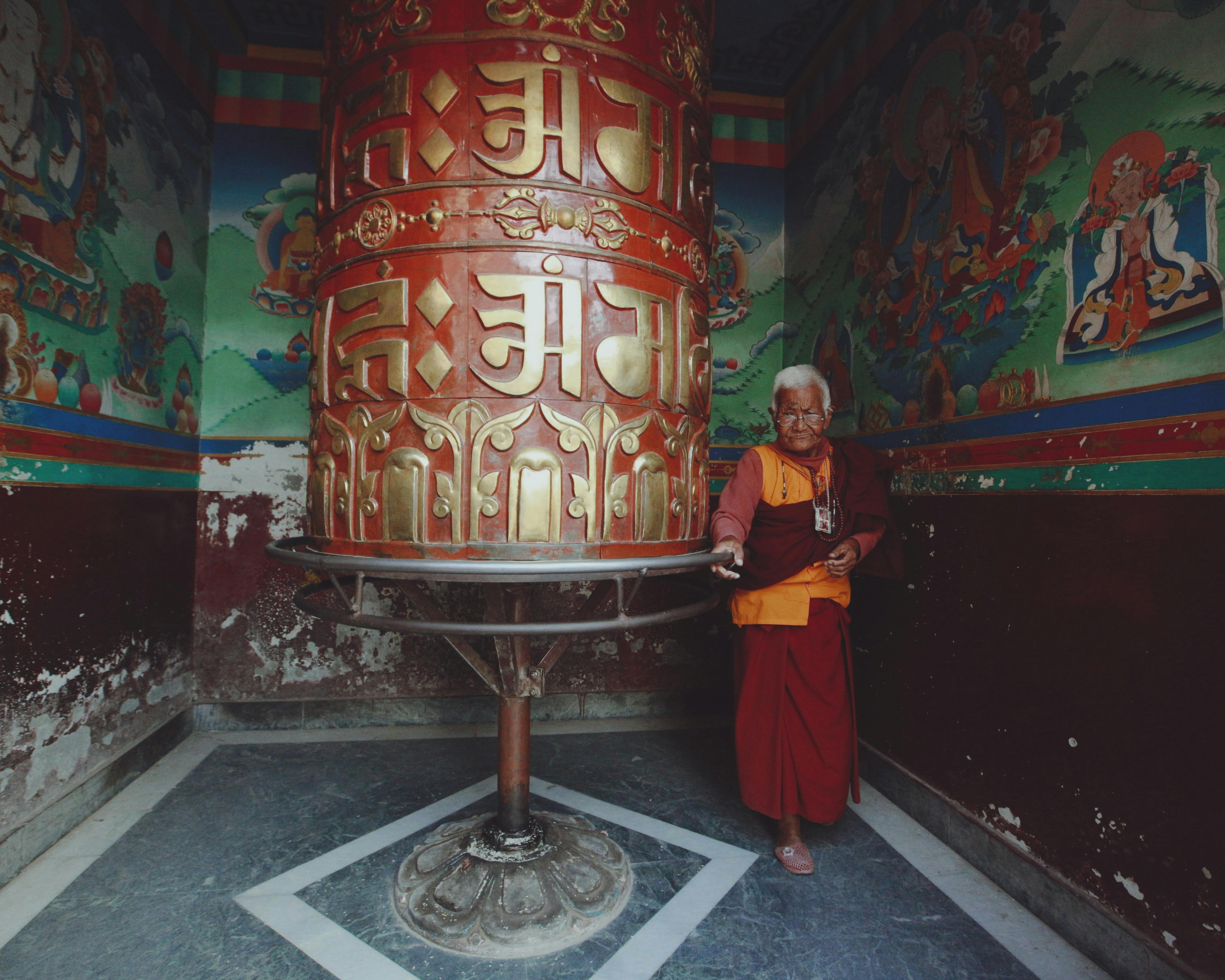 A buddhist monk stands in front of a large prayer wheel · Free Stock Photo