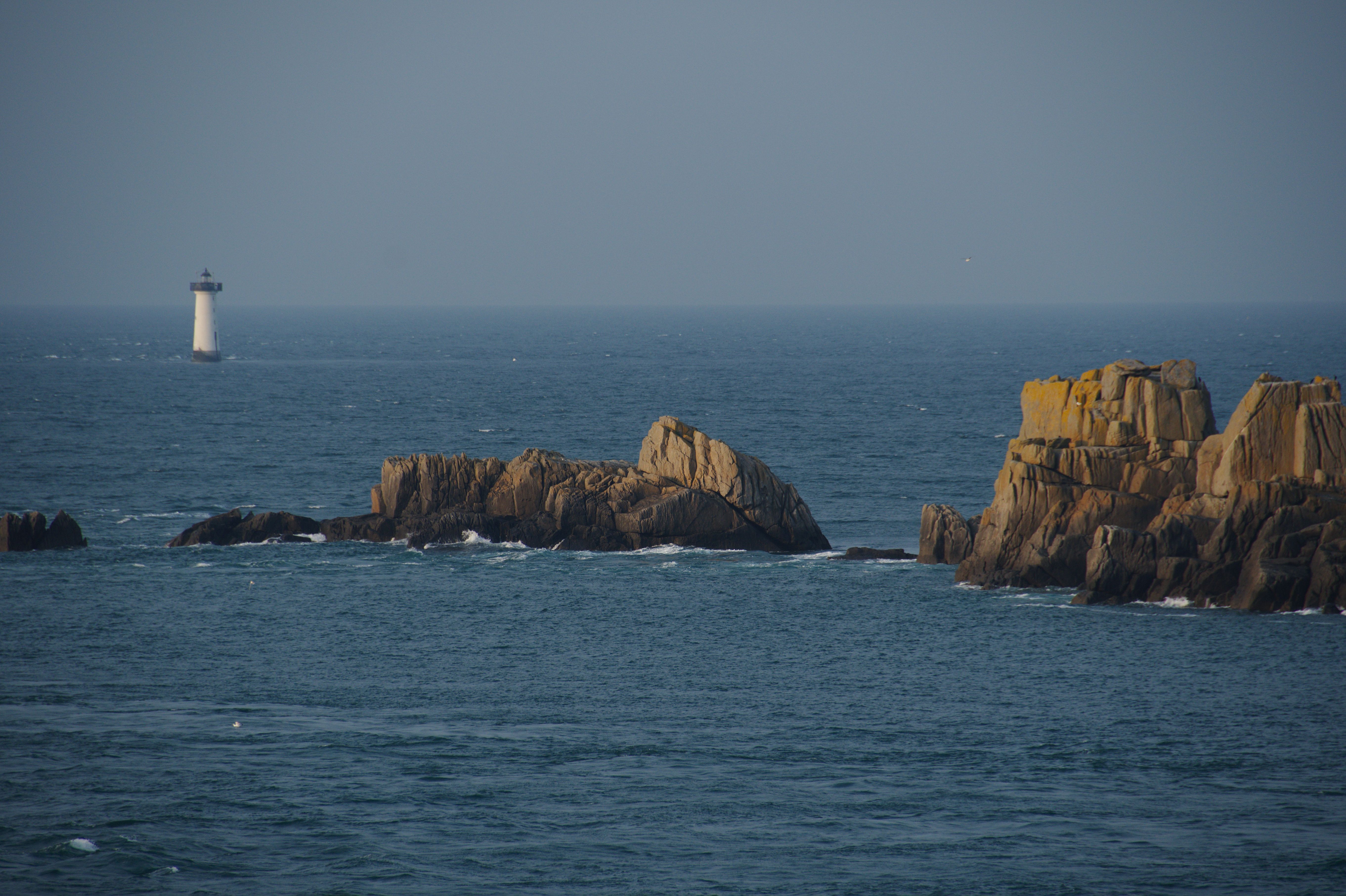 A lighthouse is in the distance with rocks in the water · Free Stock Photo
