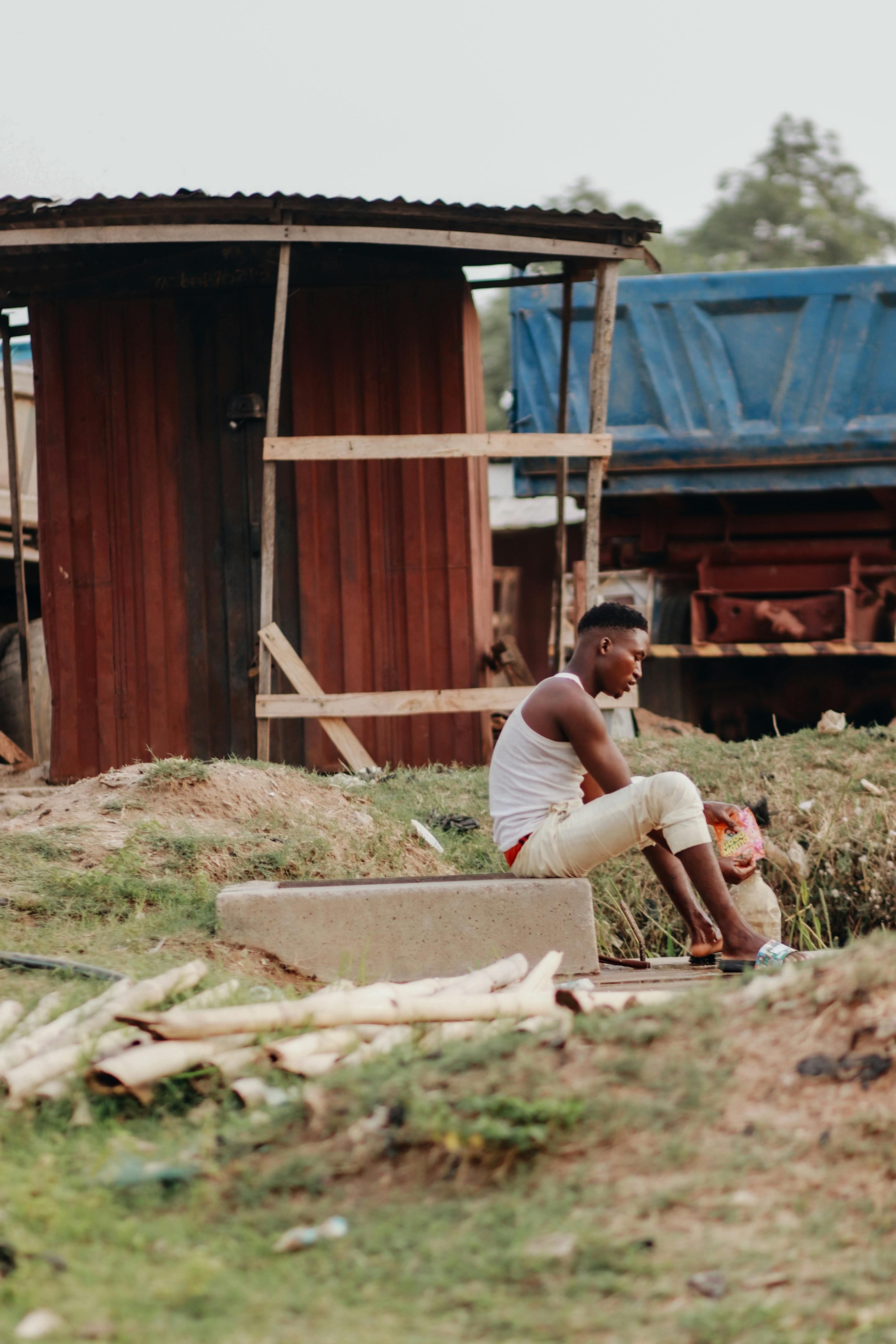 Young Man Sitting Outdoors Near Rustic Structures · Free Stock Photo