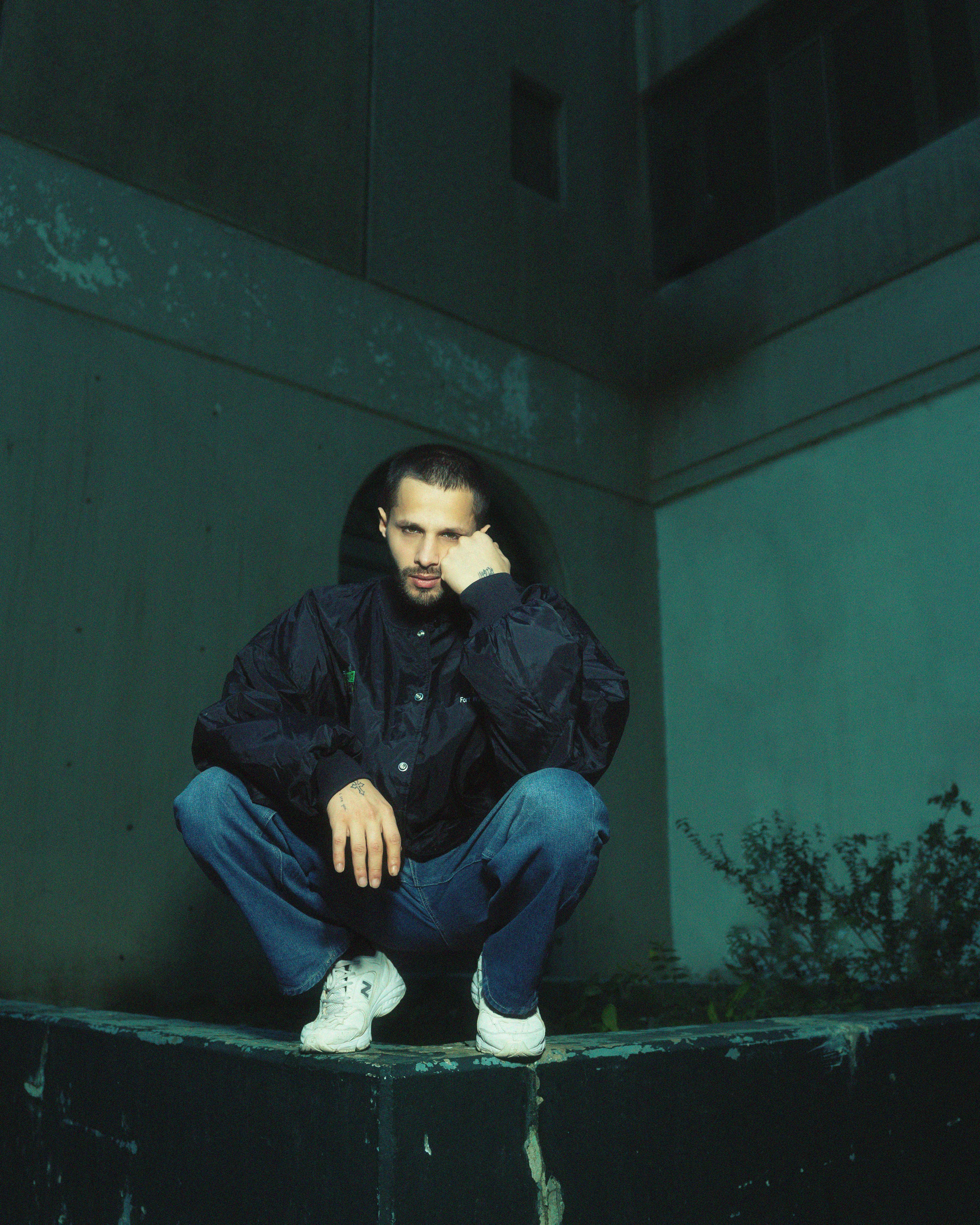 Young Man in Jeans and a Jacket Crouching on a Wall near a Building ...