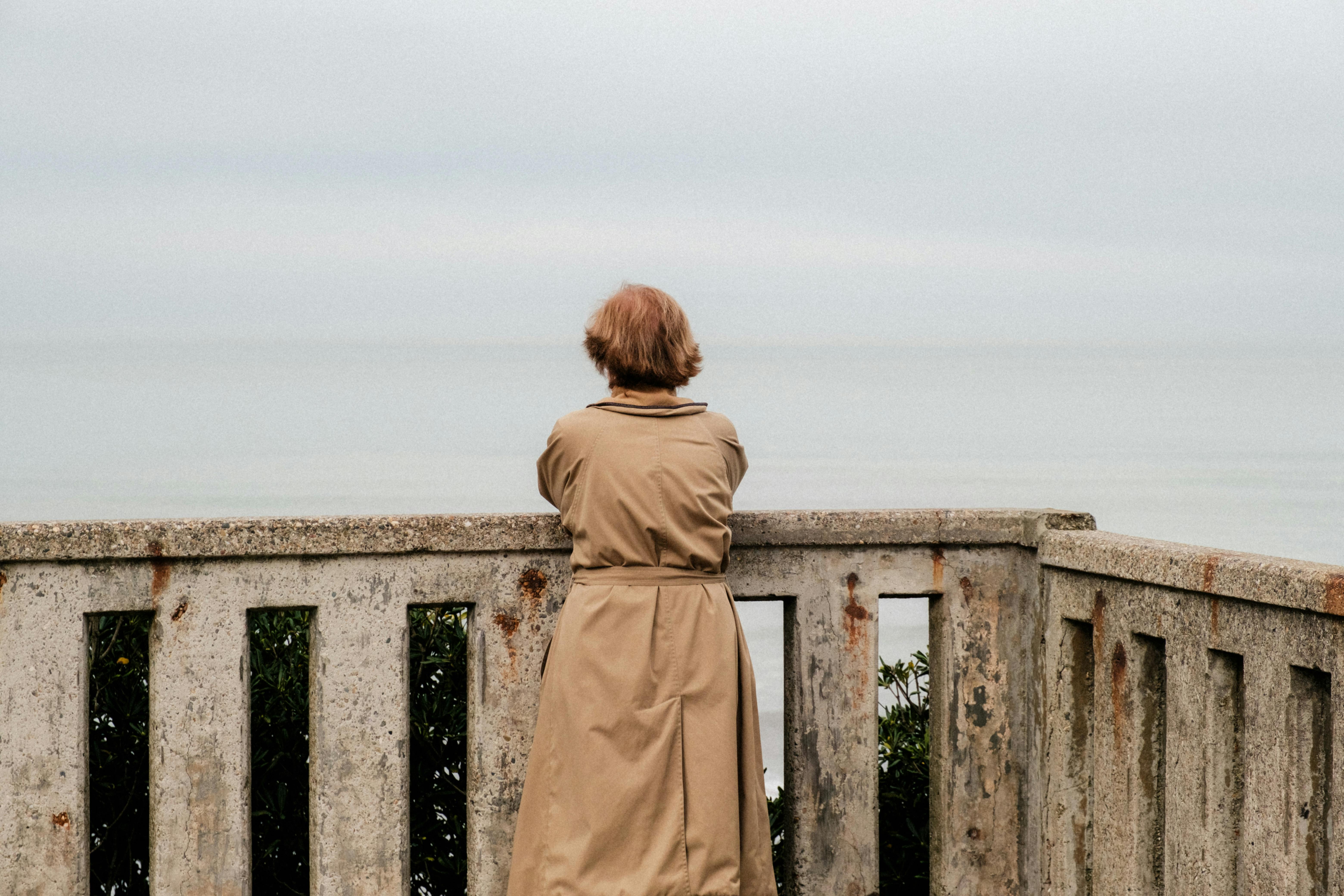 Woman in Trench Standing by Stone Railing on Sea Shore · Free Stock Photo