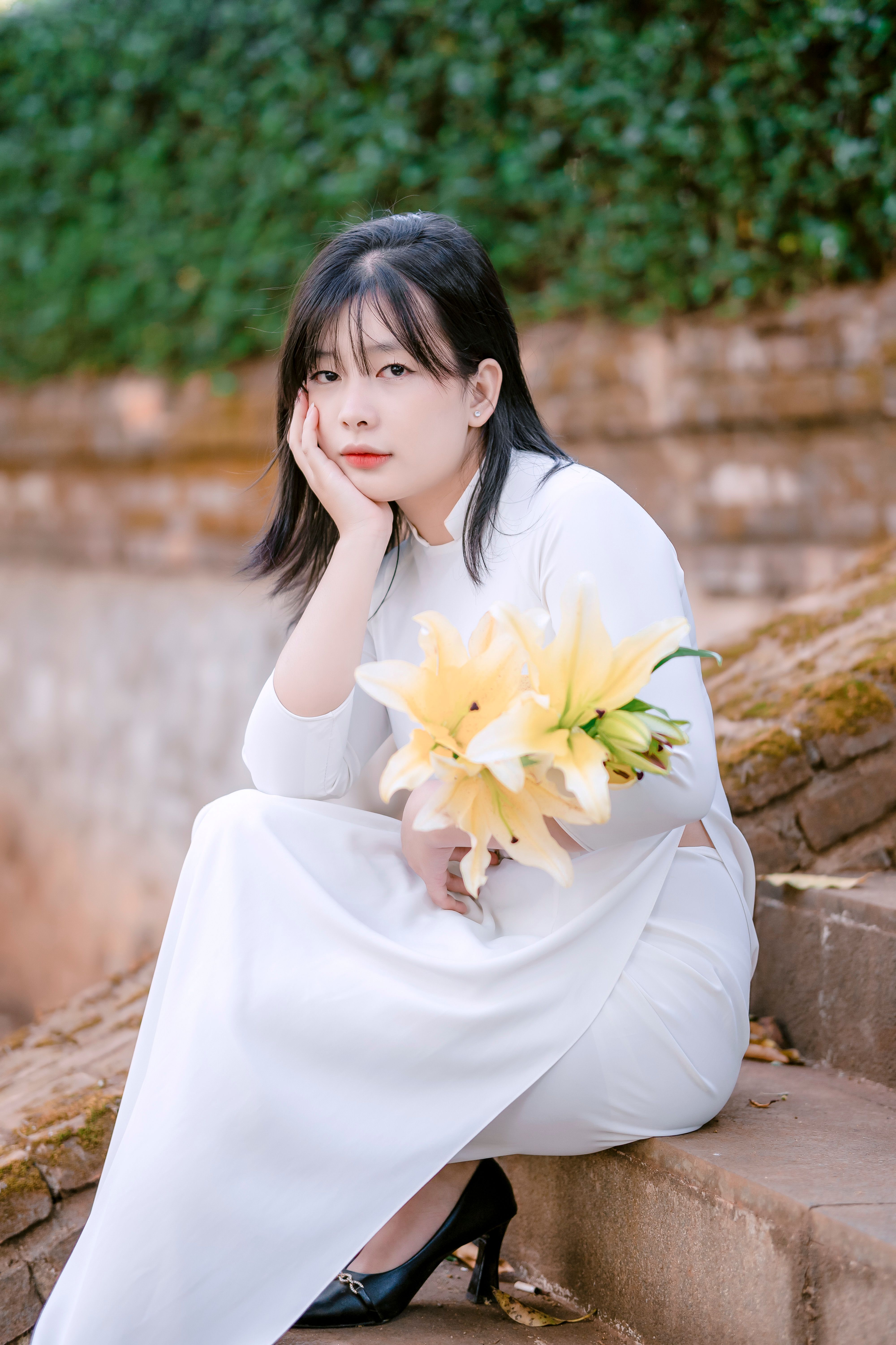 Woman in White Dress Sitting with Hand on Cheek and Holding Flowers ...