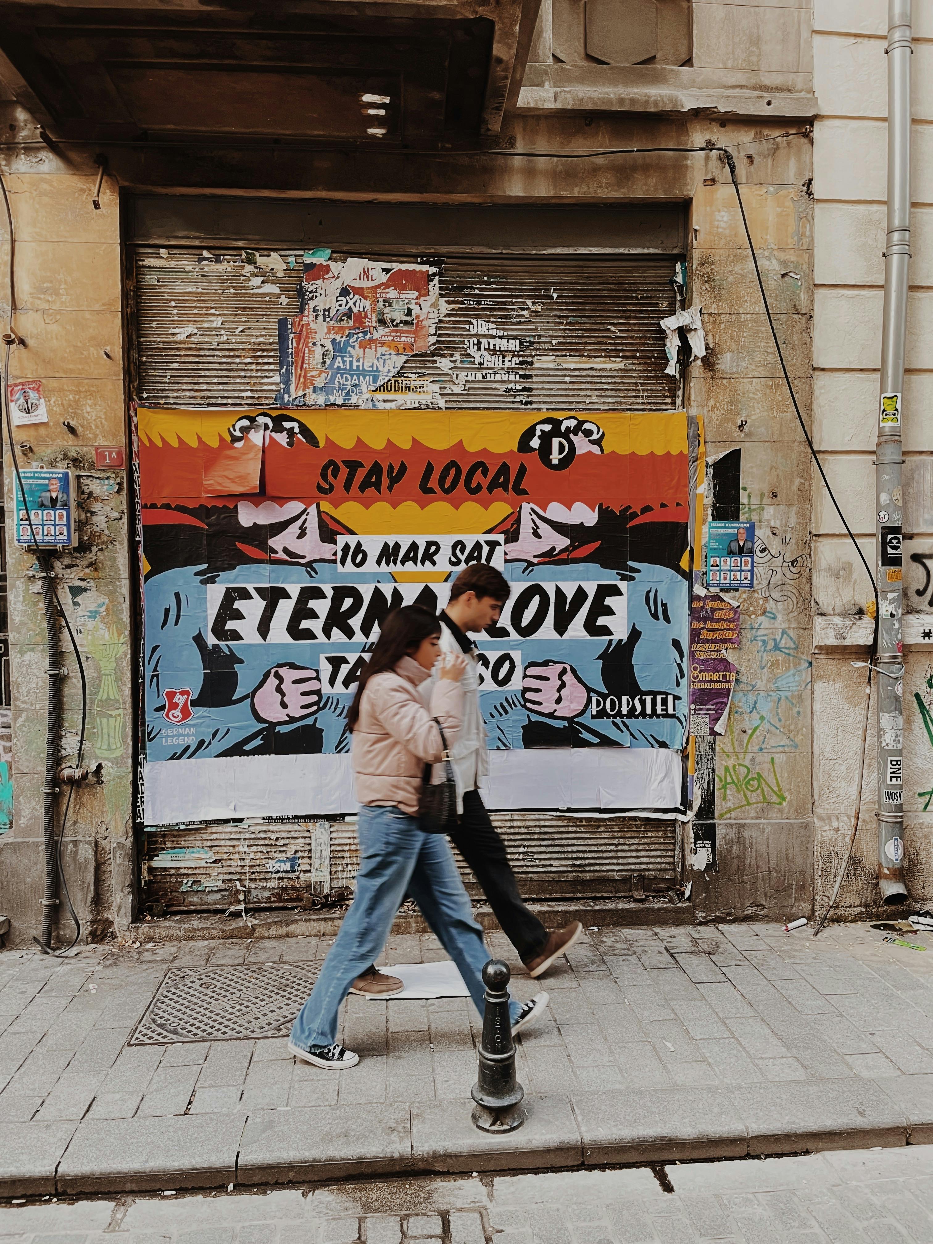 Woman and Man Walking near Poster on Wall · Free Stock Photo