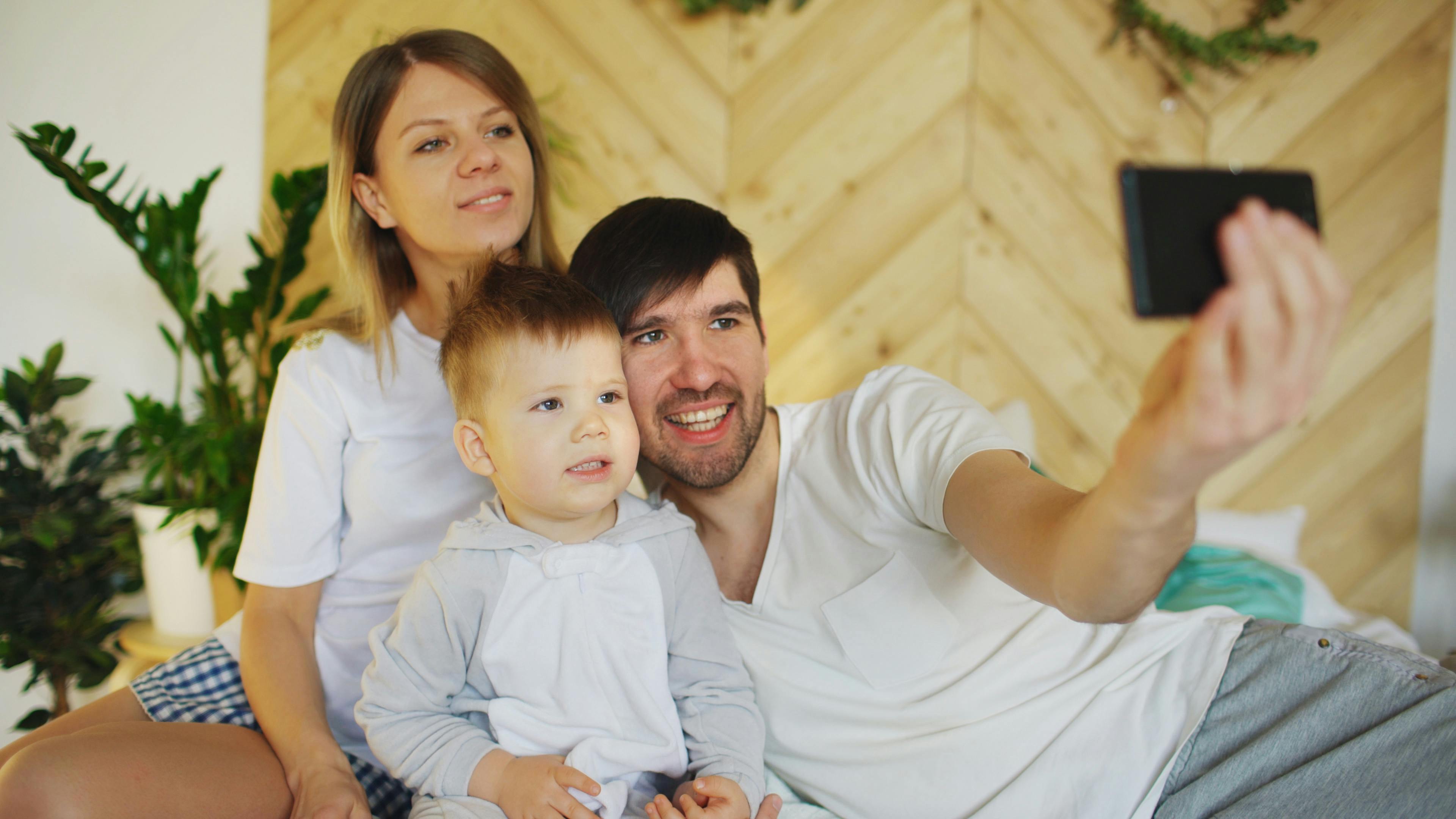 Smiling parents with baby taking selfie family photo on bed at home ...