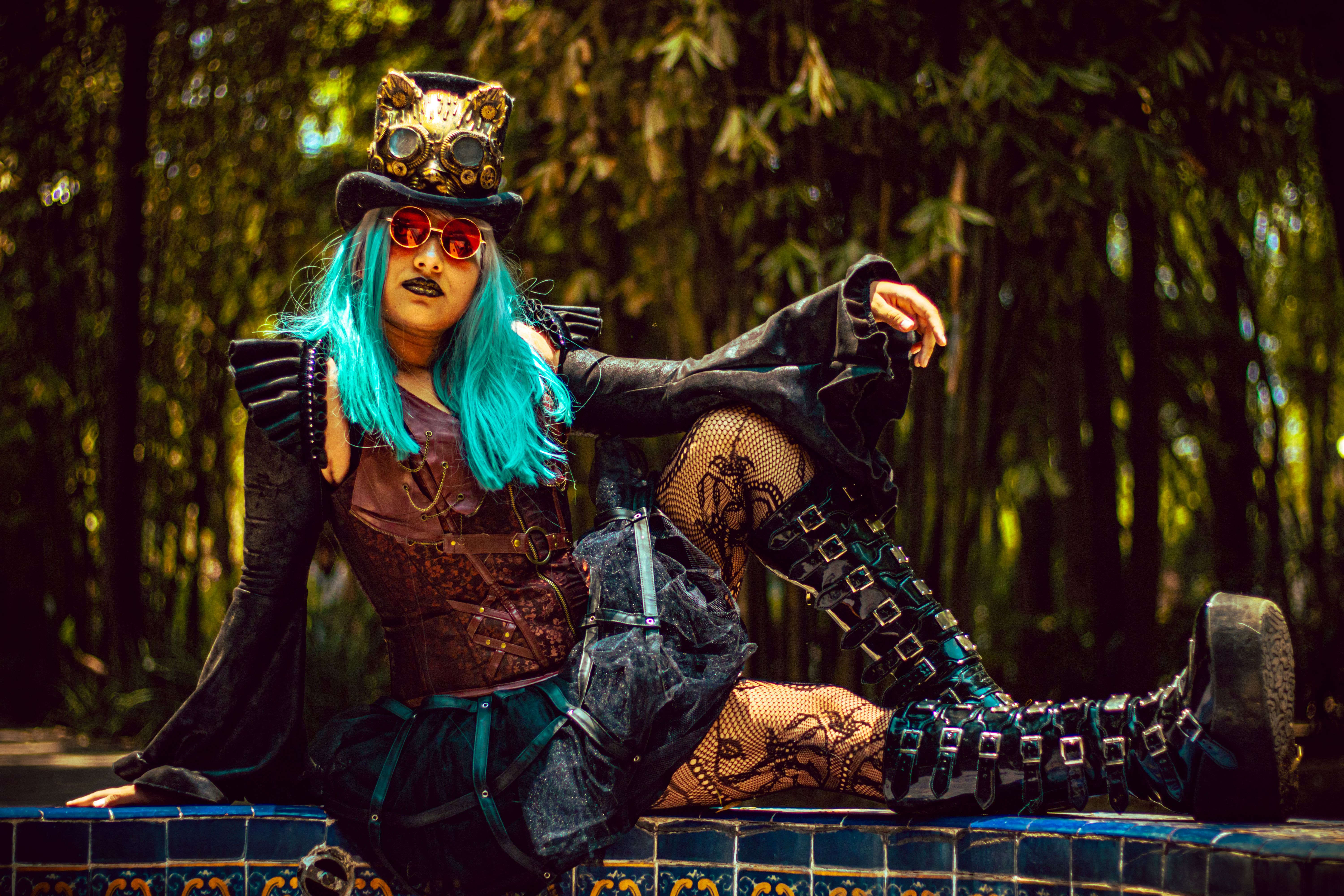 A woman with blue hair and a steampunk costume sitting on a bench ...