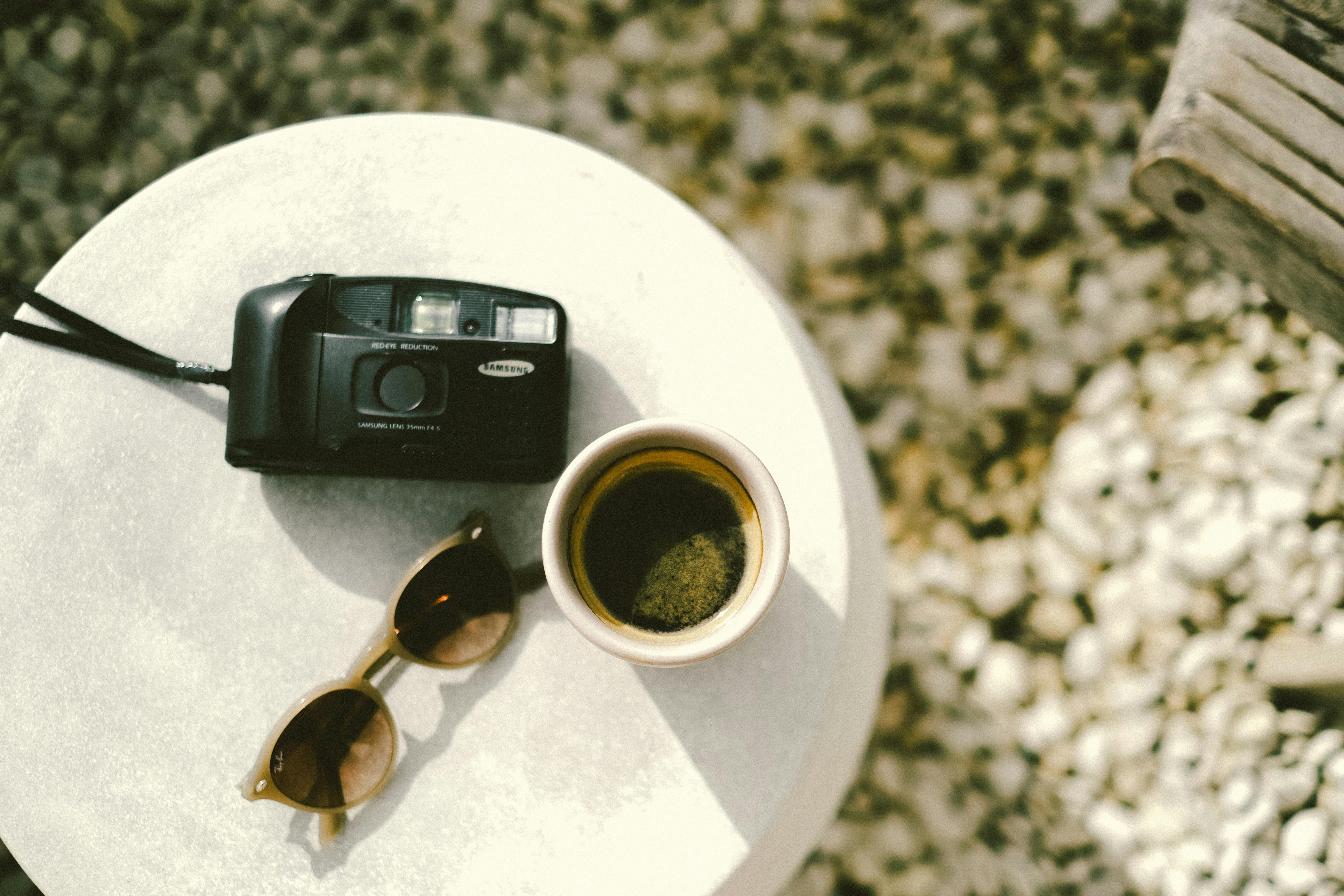 A camera and sunglasses on a table next to a cup of coffee · Free Stock ...