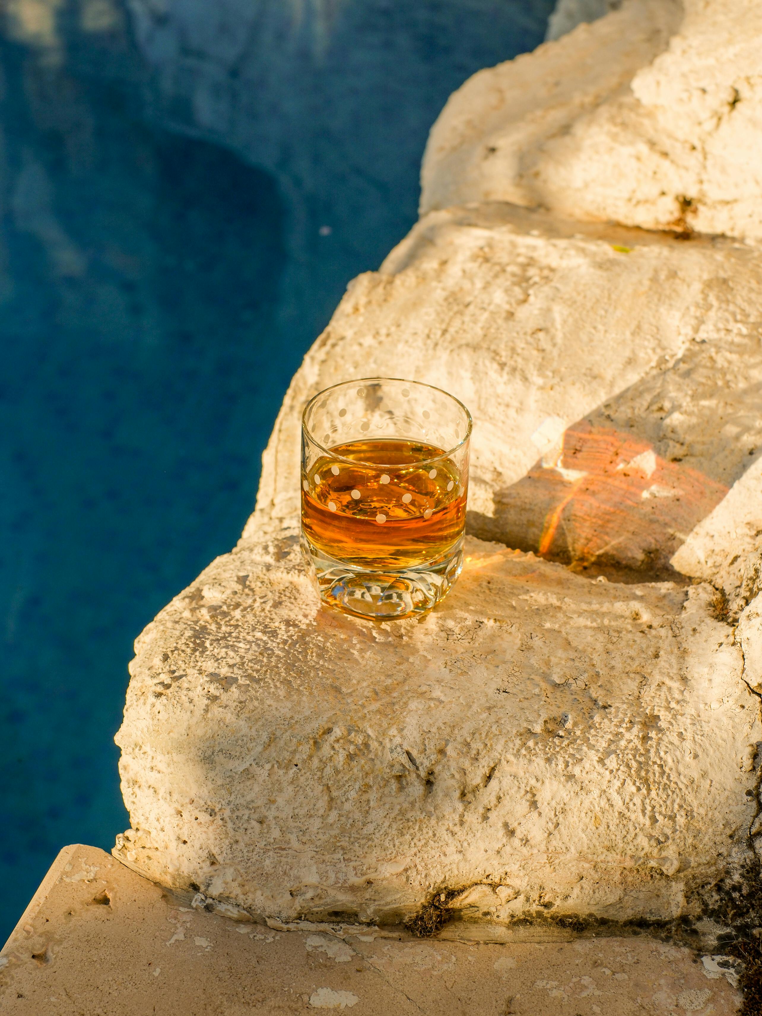 A glass of whiskey sitting on a rock by a pool · Free Stock Photo