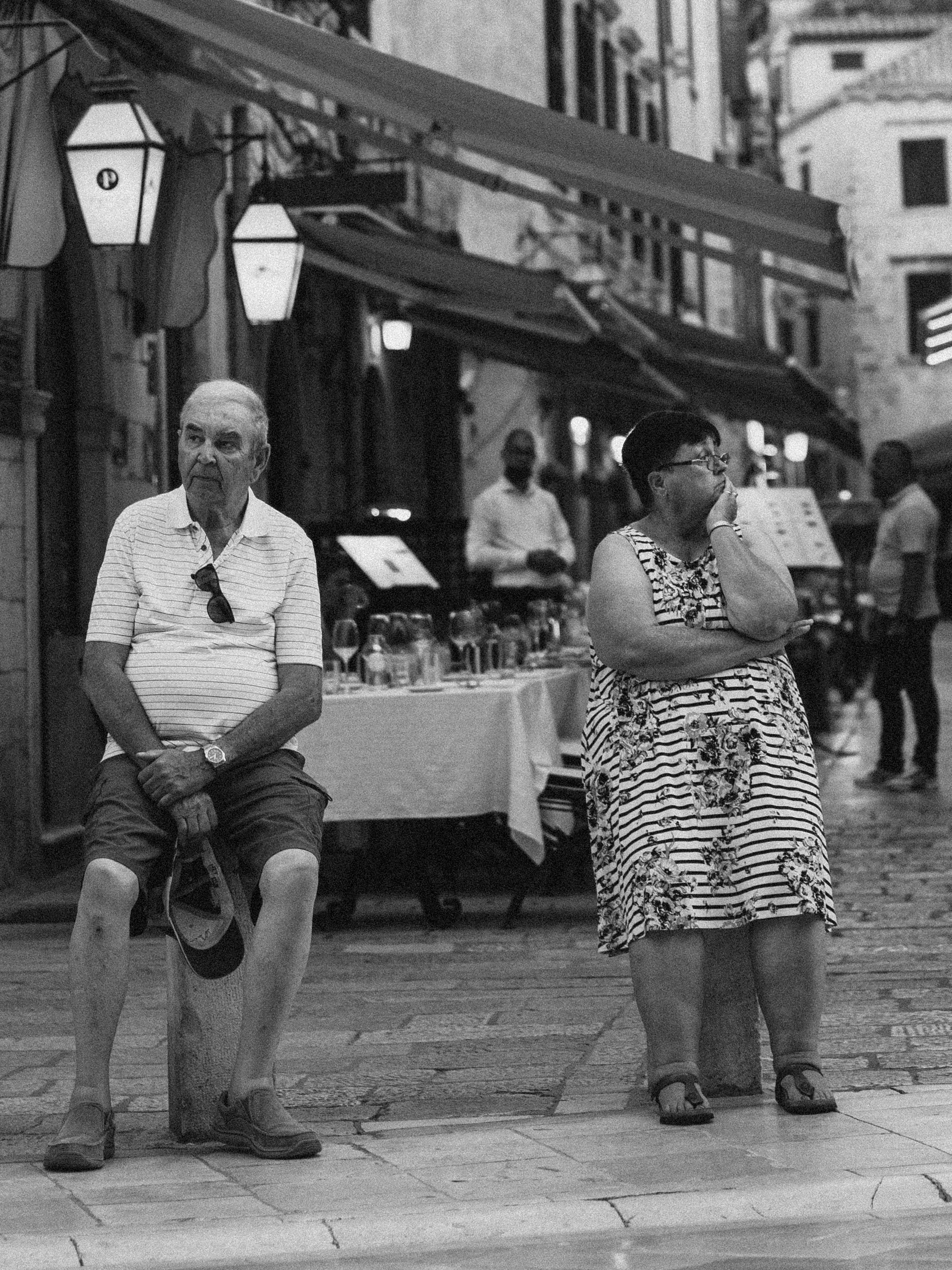 Elderly Man and Woman Sitting on Posts on Sidewalk · Free Stock Photo