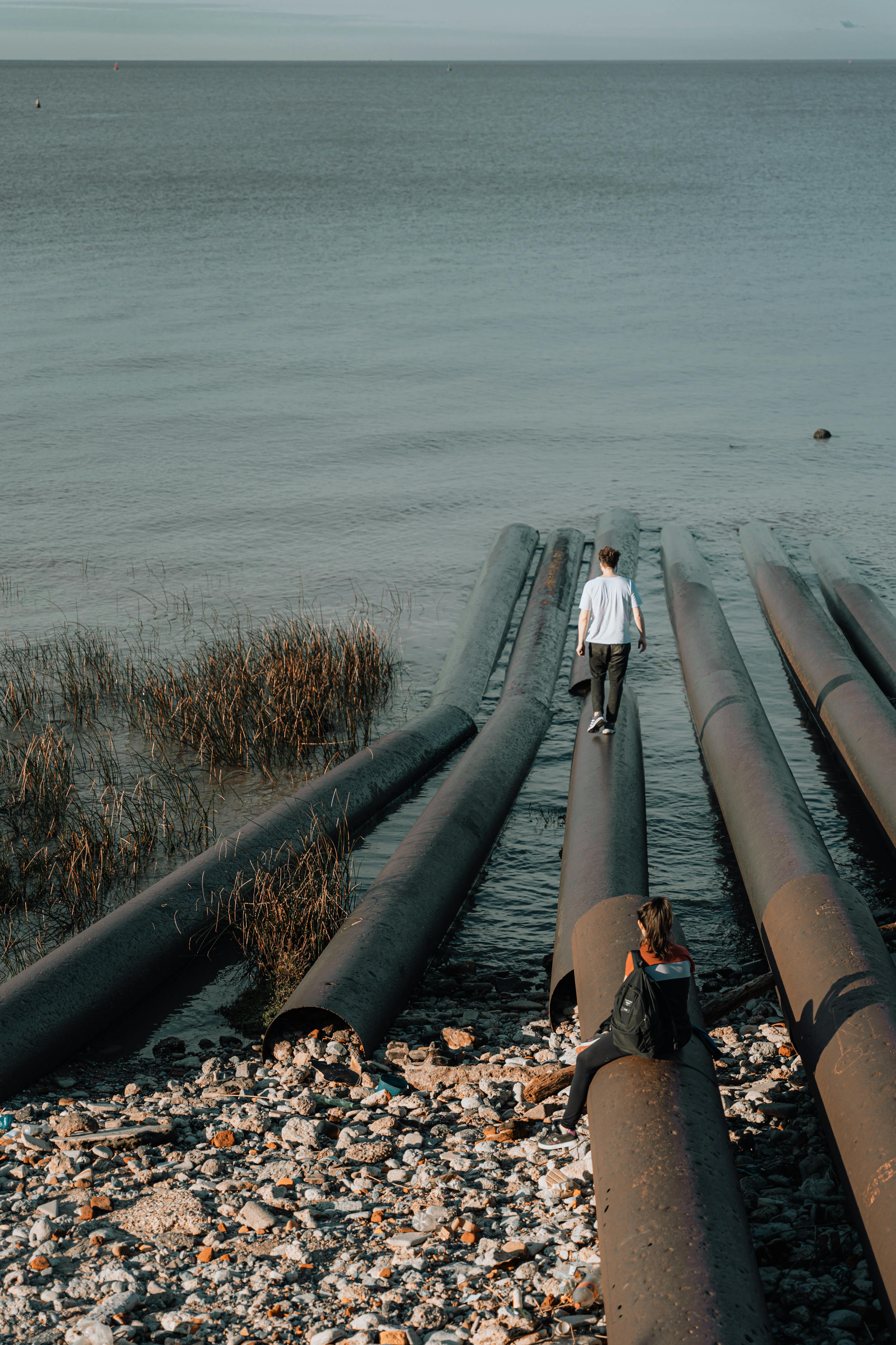 Woman Sitting and Man Walking on Pipes on Sea Shore · Free Stock Photo