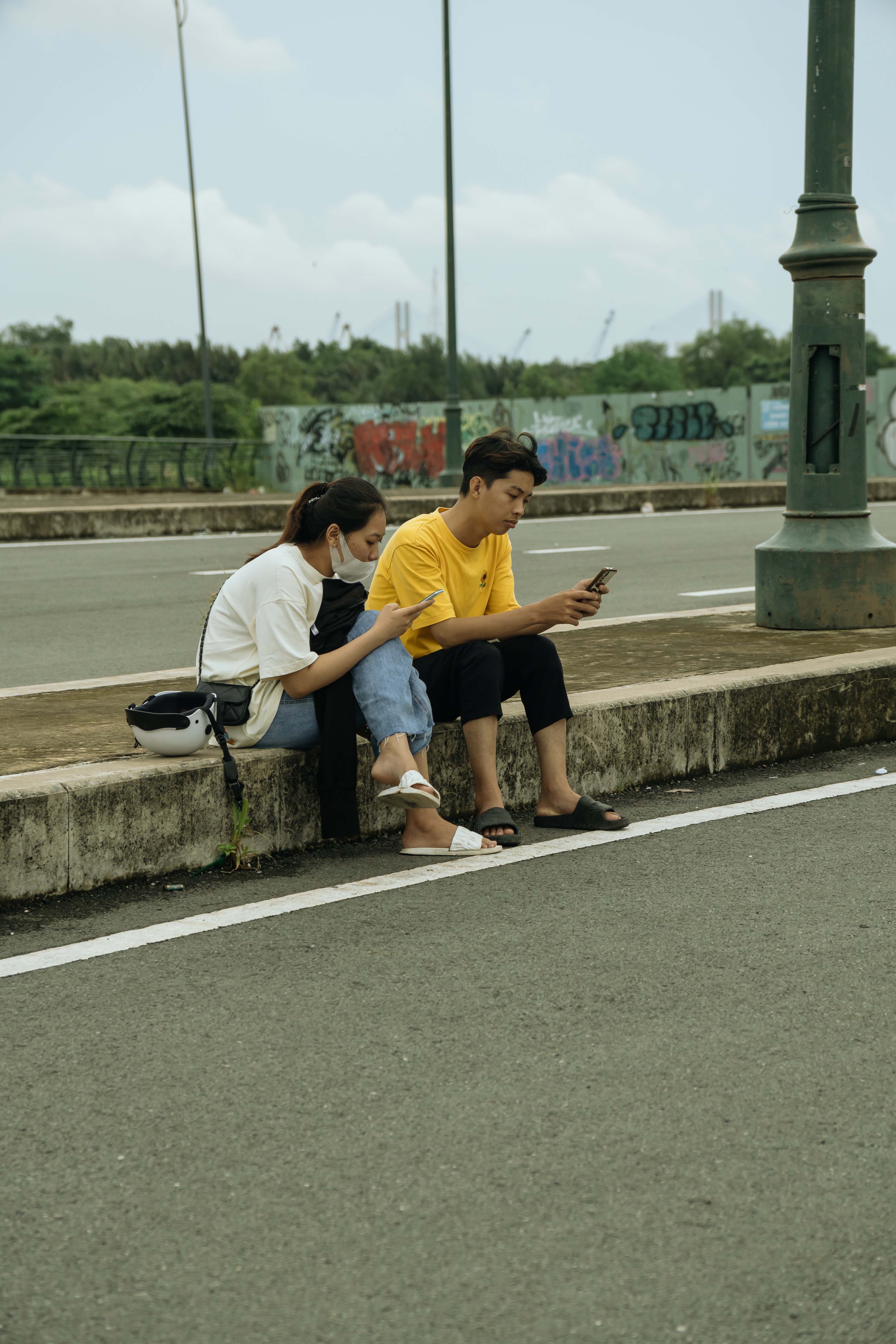 Two people sitting on the side of the road looking at their cell phones ...