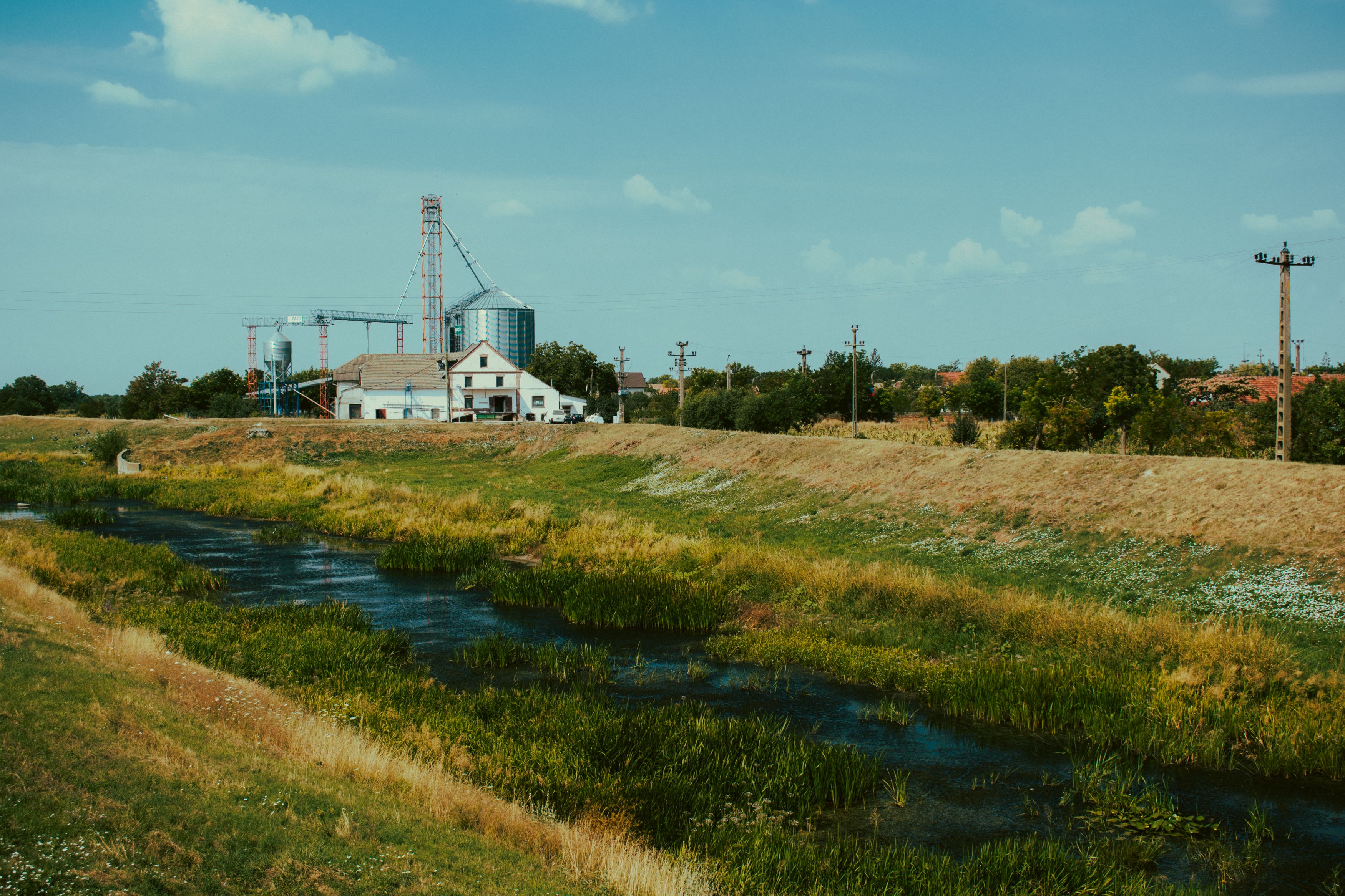 A river runs through a field with a farm · Free Stock Photo