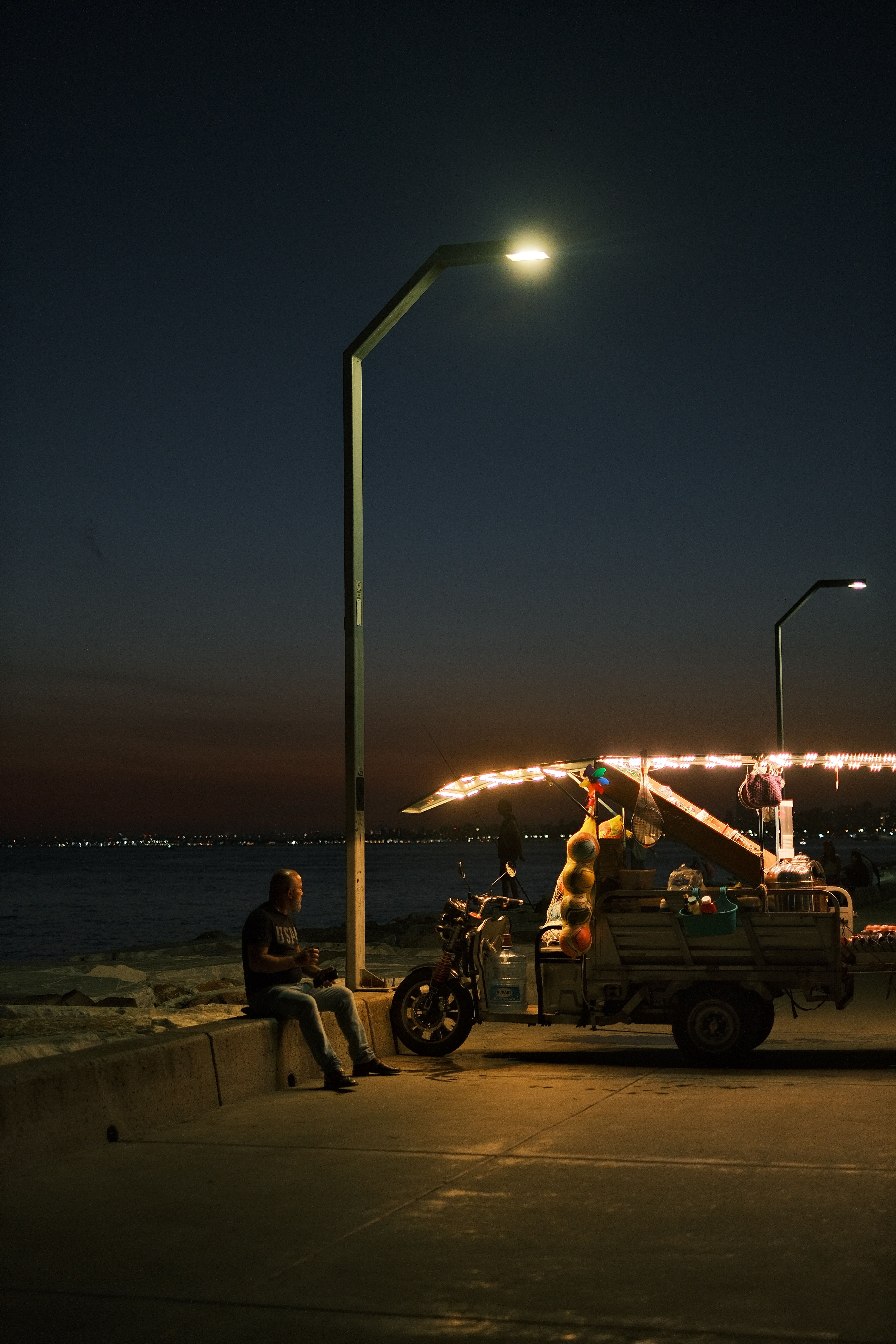 A man sitting on a bench next to a food cart at night · Free Stock Photo