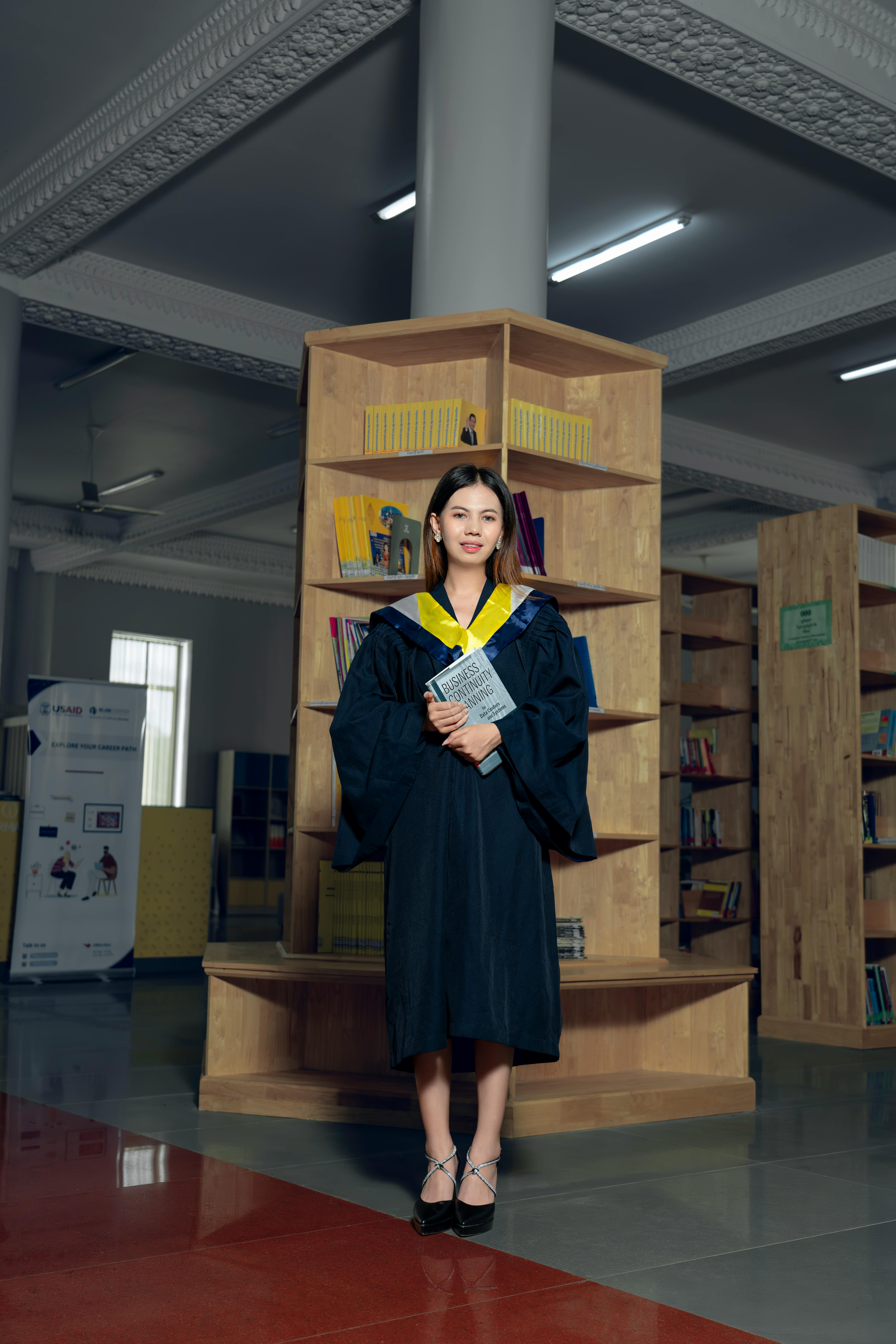 A woman in a graduation gown standing in front of a bookcase · Free ...