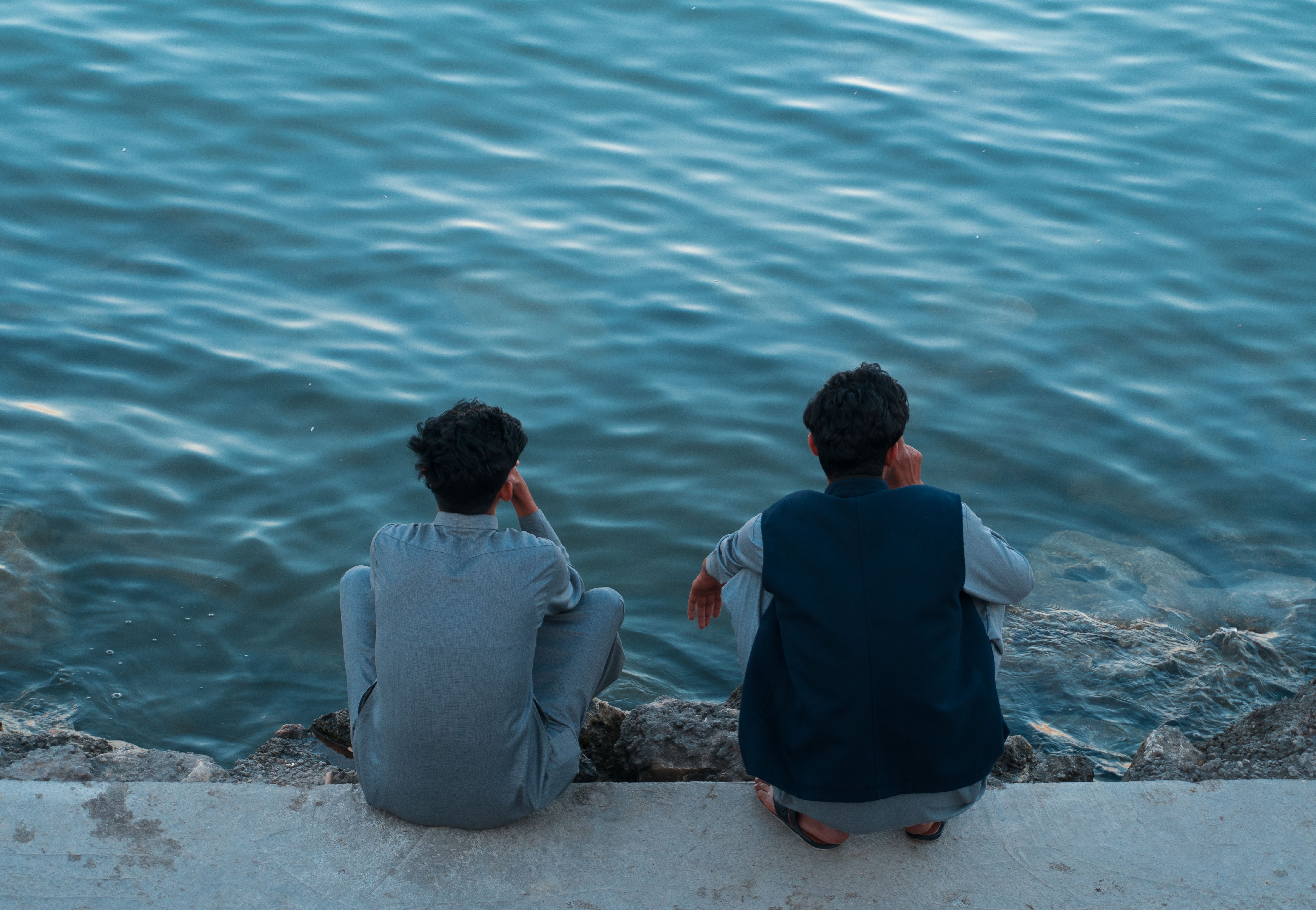 Two men sitting on the edge of a body of water · Free Stock Photo