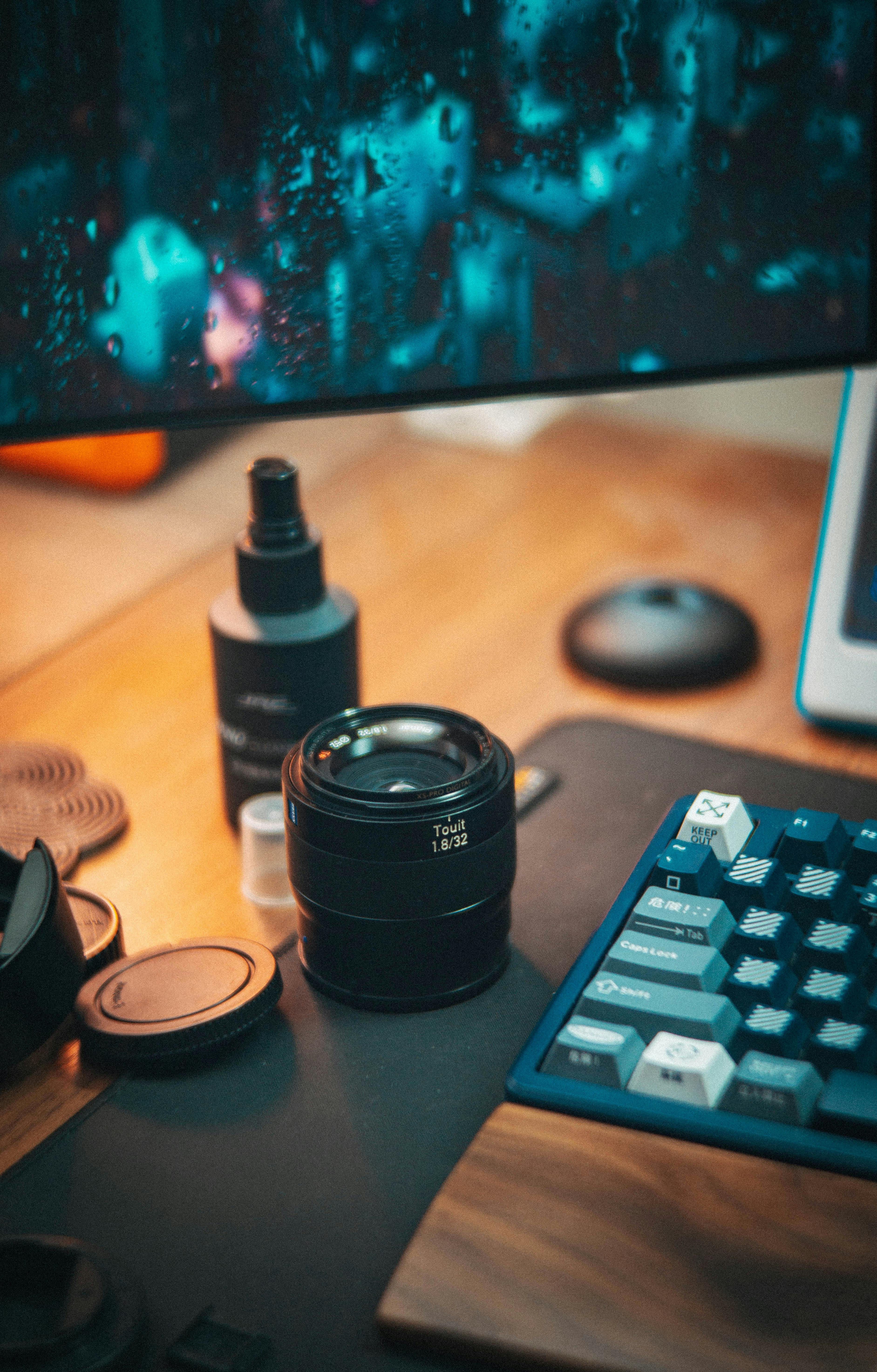 A computer monitor, camera lens and keyboard on a desk · Free Stock Photo