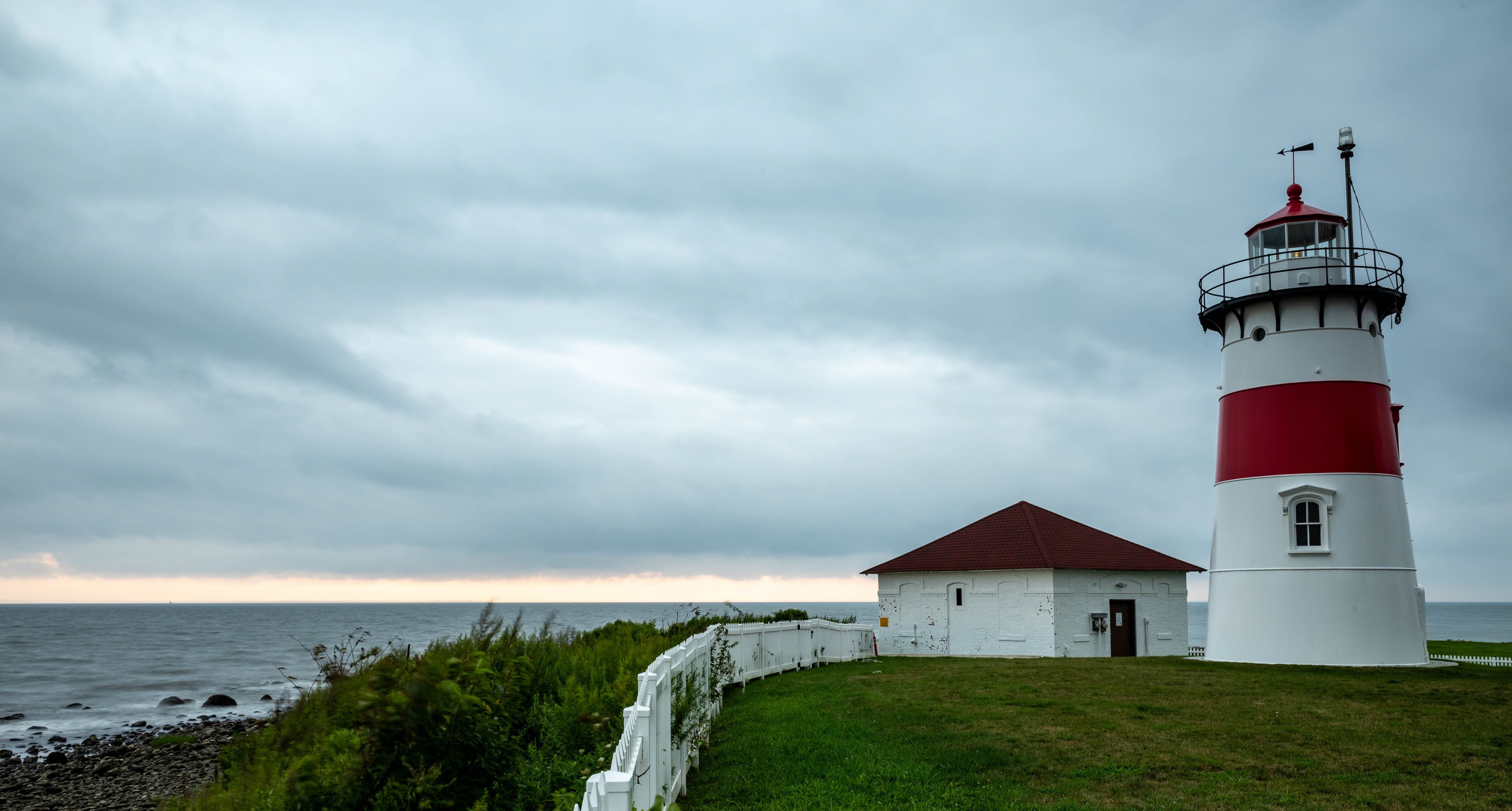 A lighthouse sits on a grassy hill next to the ocean · Free Stock Photo