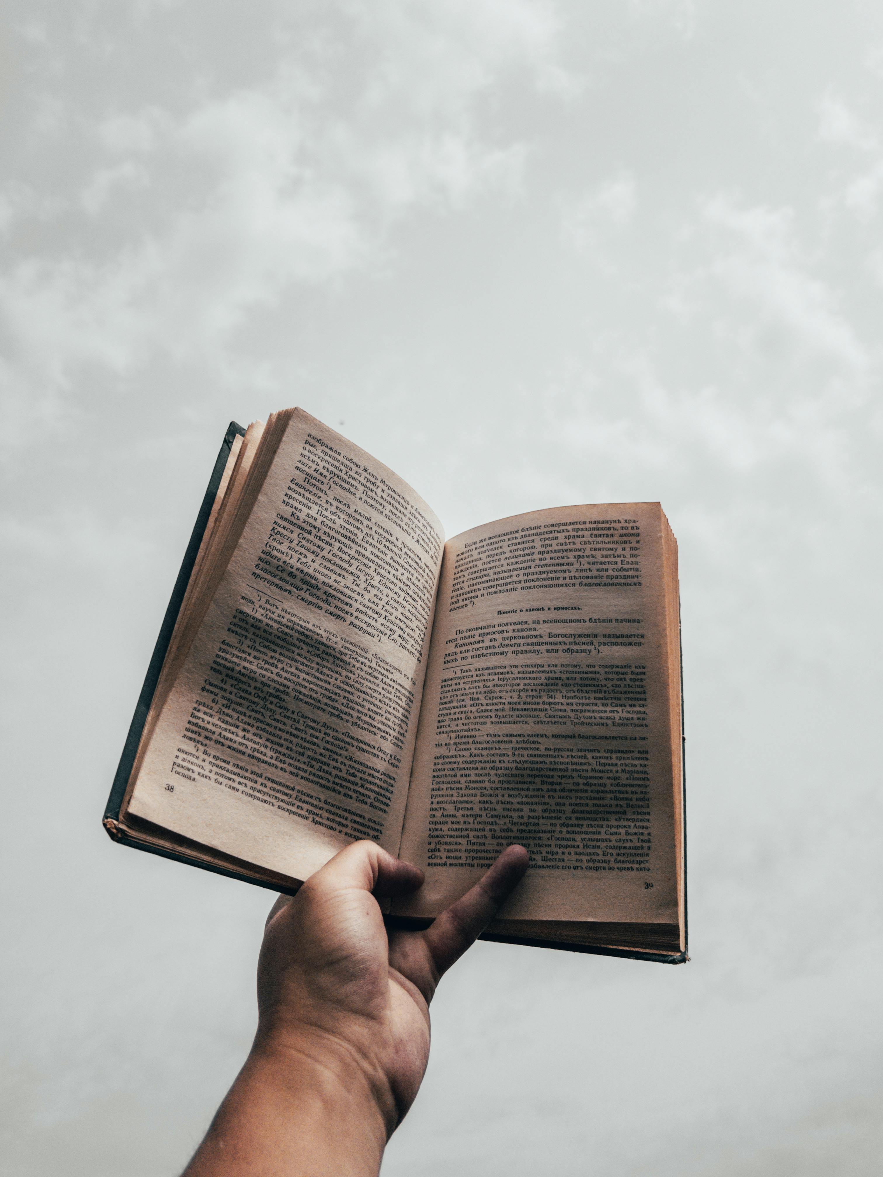 A person holding an open book in front of a cloudy sky · Free Stock Photo