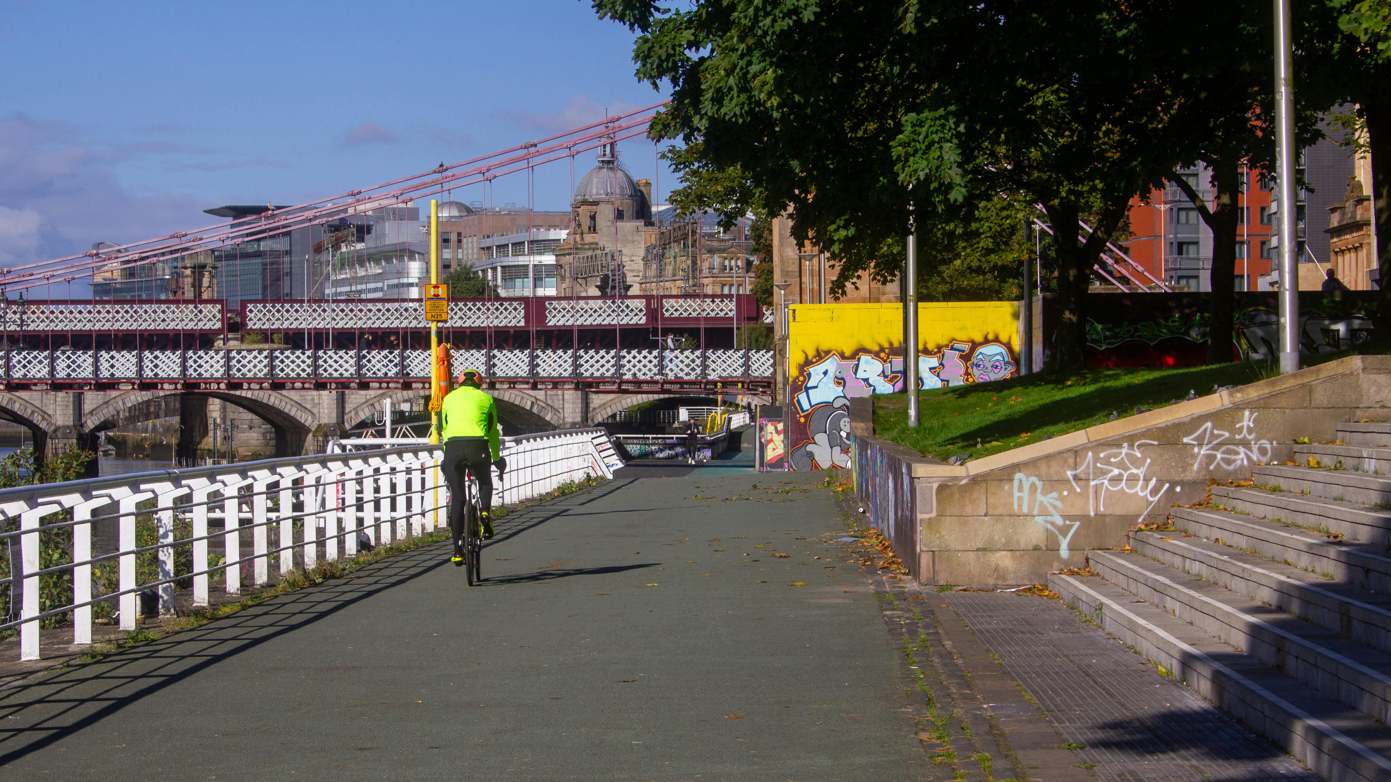 Cyclist on Riverfront Pathway with Bridge and Graffiti · Free Stock Photo
