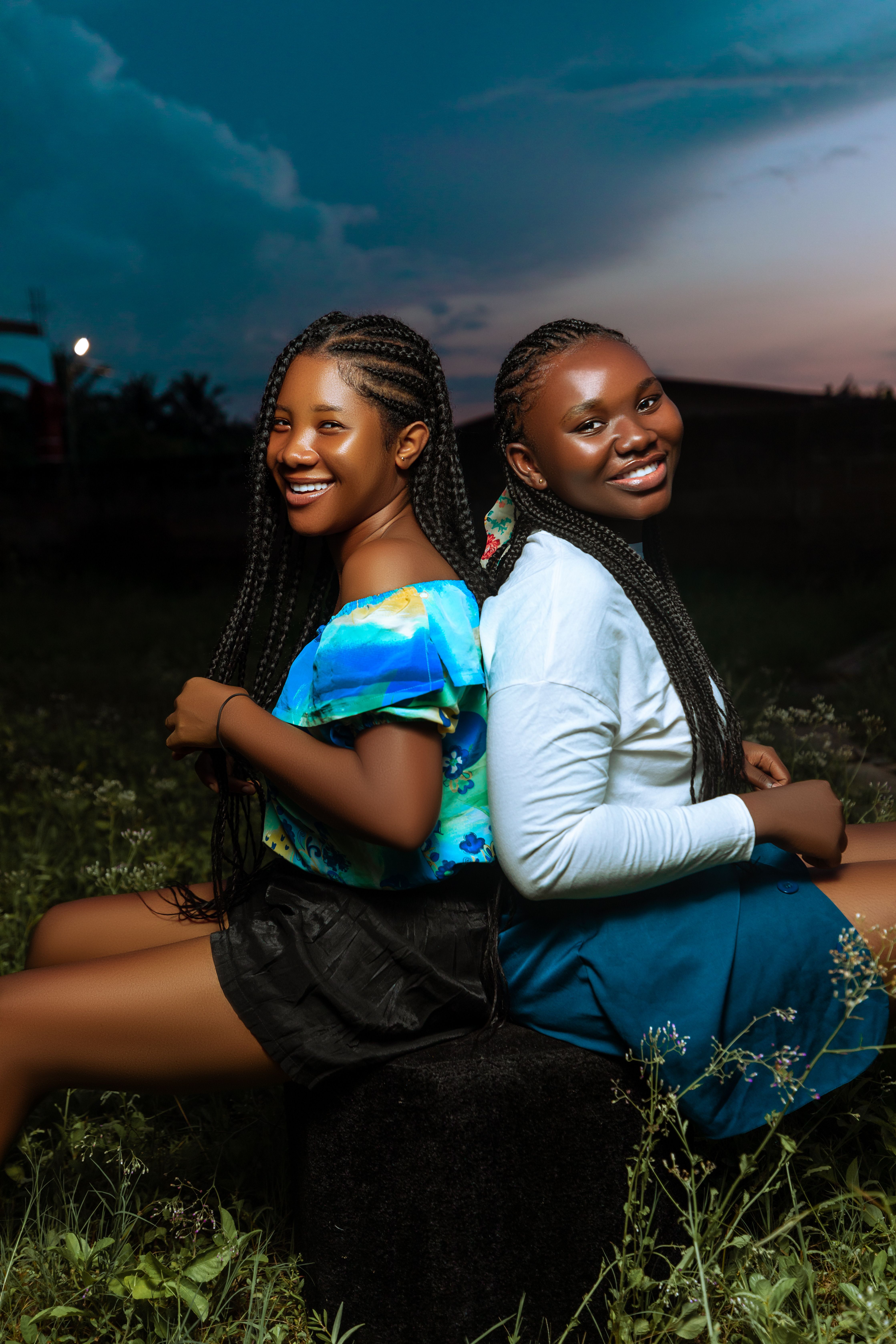 Two Smiling Girls Sitting Back-to-Back at Dusk · Free Stock Photo