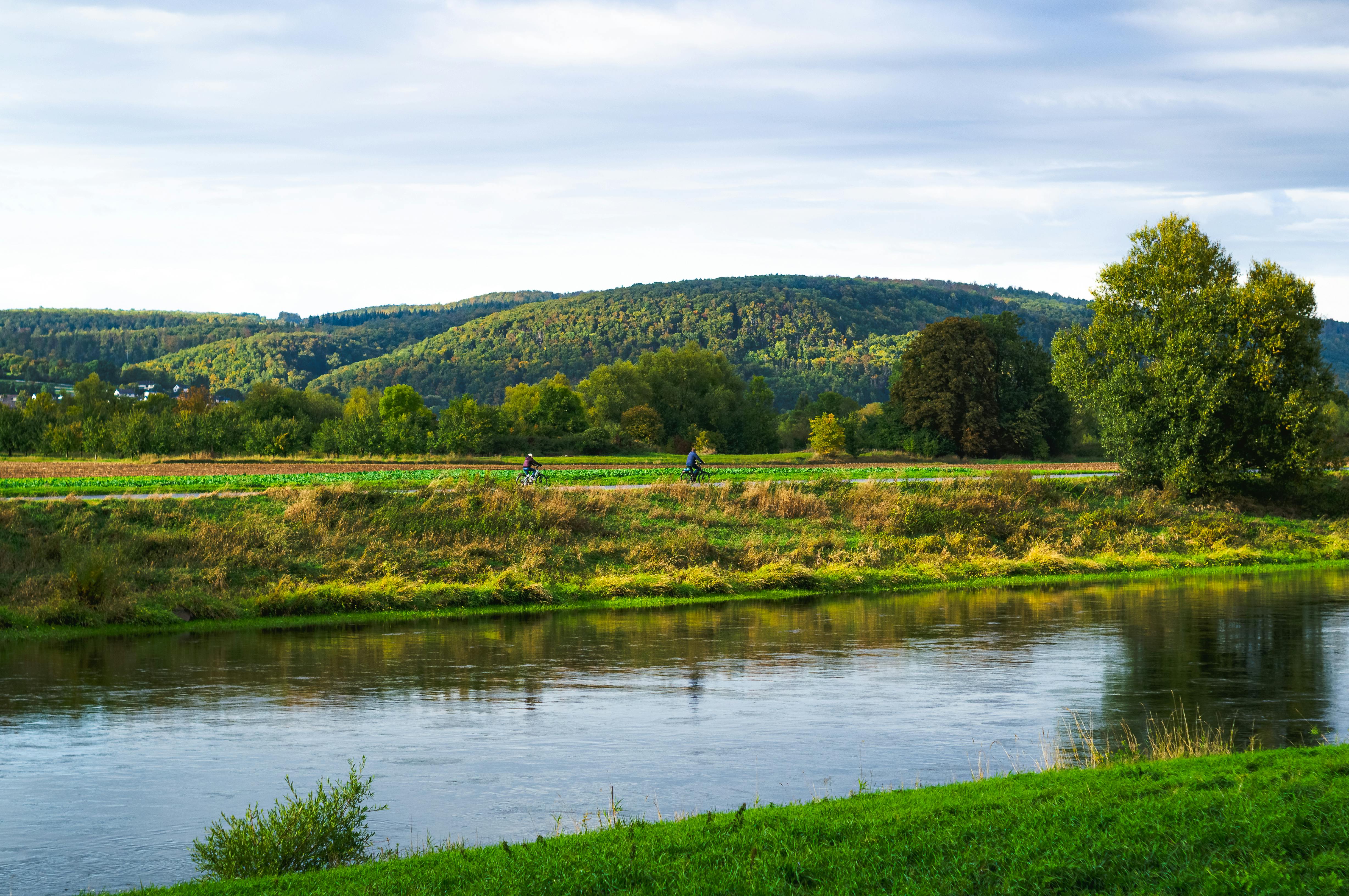 Scenic Riverbank with Cyclists in Lush Landscape · Free Stock Photo