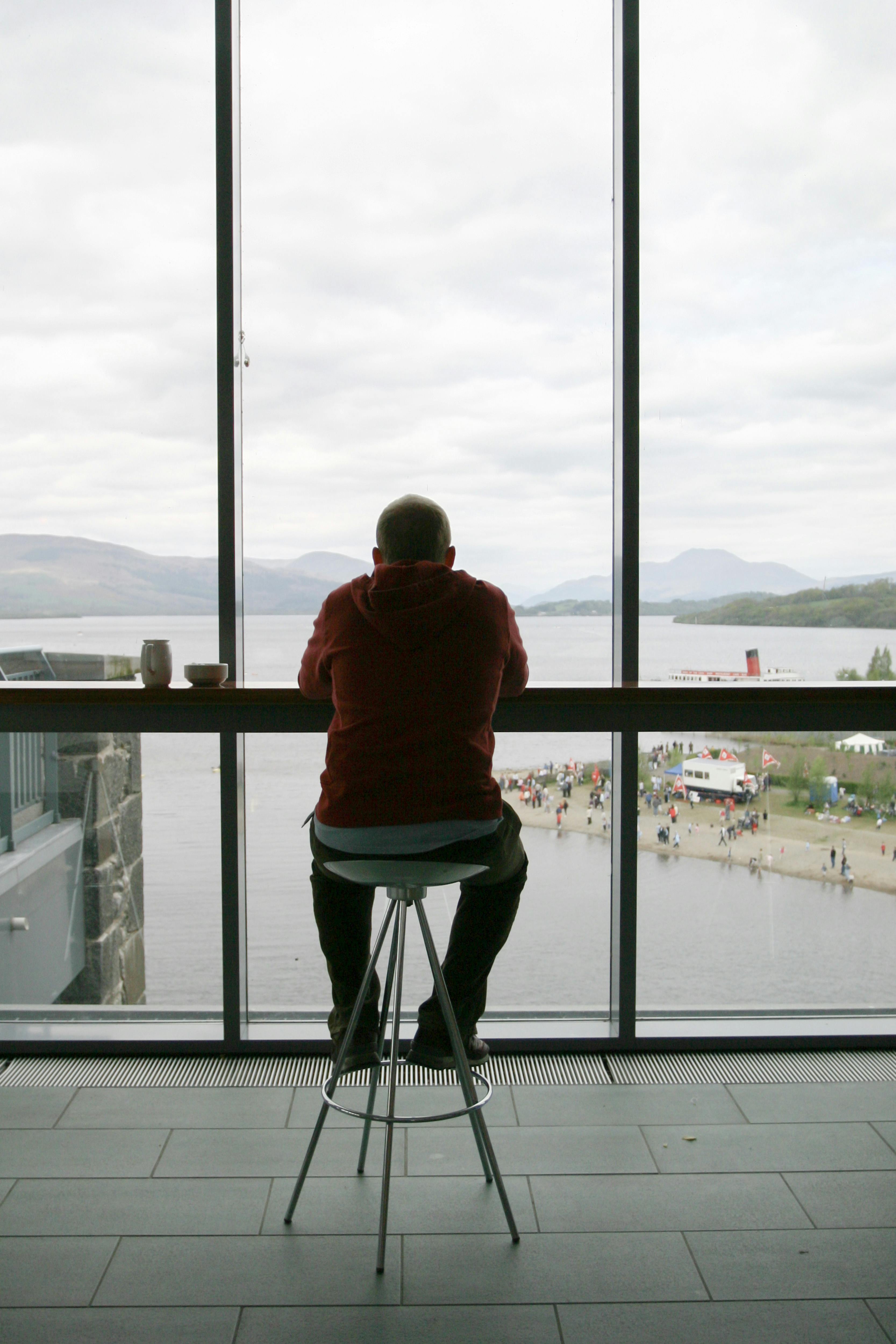 Man Sitting by Large Window Overlooking Lake · Free Stock Photo