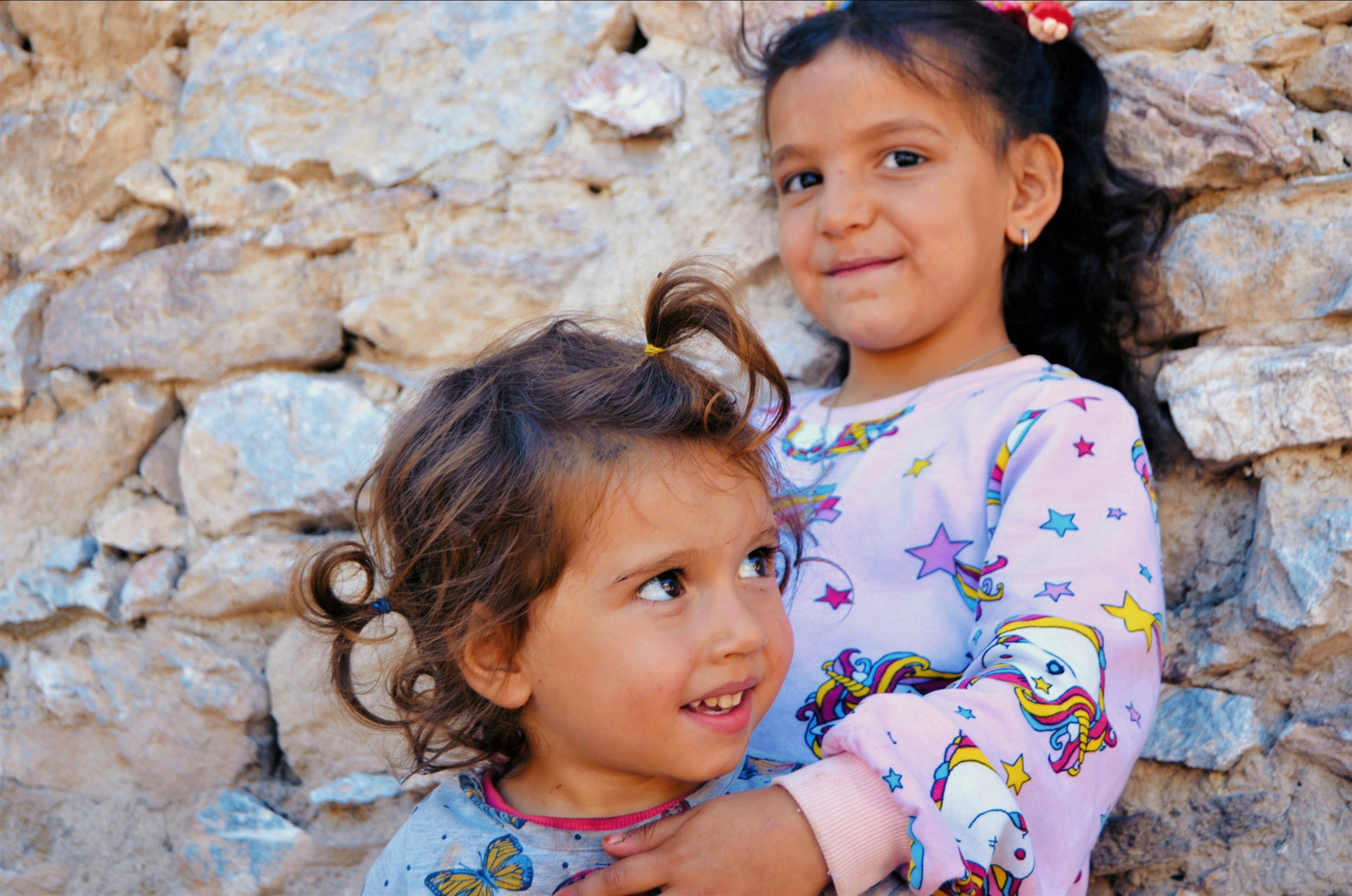 Two Smiling Children in Colorful Outfits by Stone Wall · Free Stock Photo